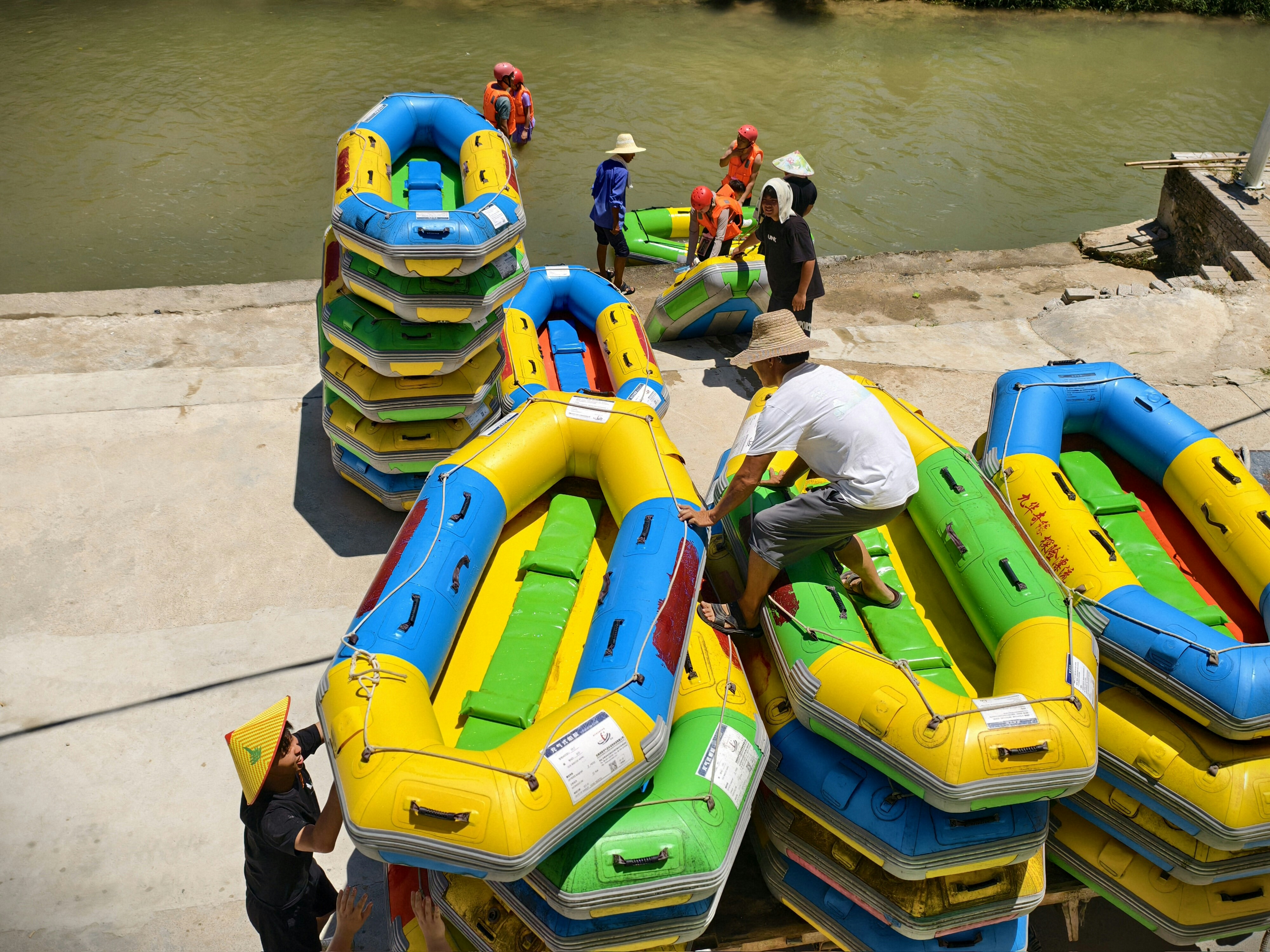 Vibrant inflatable boats stacked by the riverside, with people guiding and inspecting them near a calm waterway.