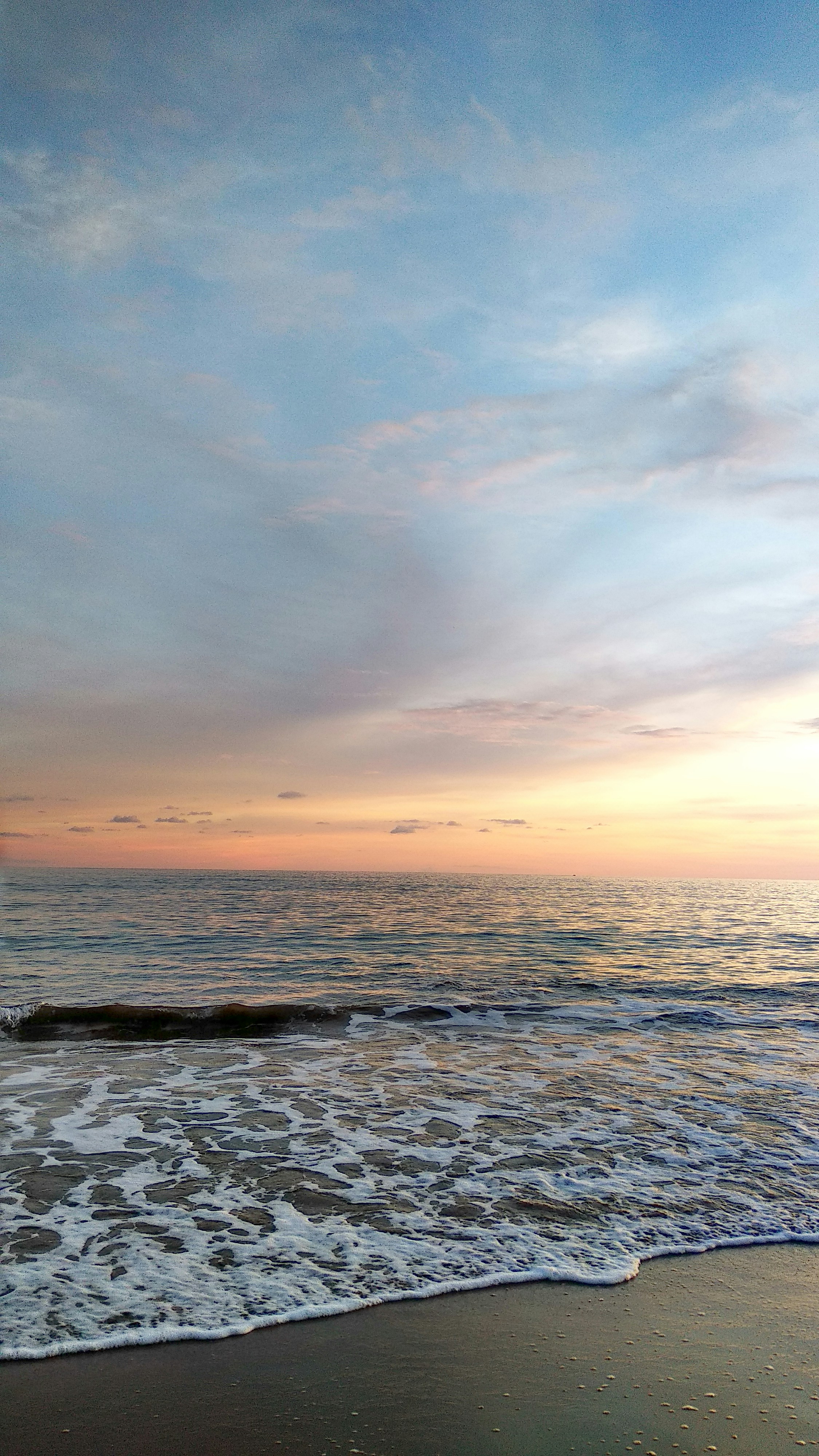 Sunset-lit horizon over calm sea with foamy shoreline and gentle waves; a tranquil coastal scene.
