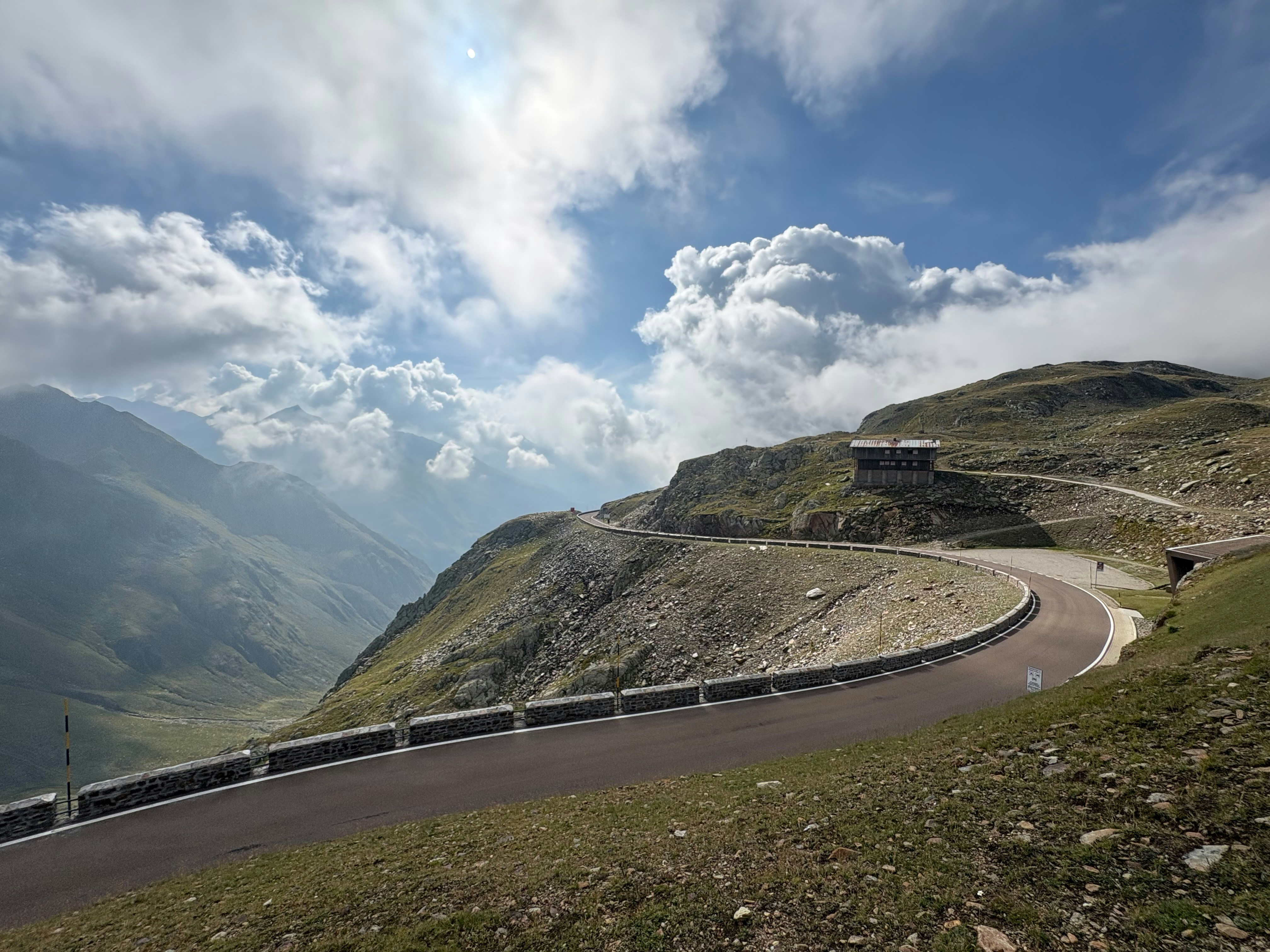 Winding mountain road hugging a grassy ridge under dramatic clouds; a distant guardrail and hillside add depth and scale.