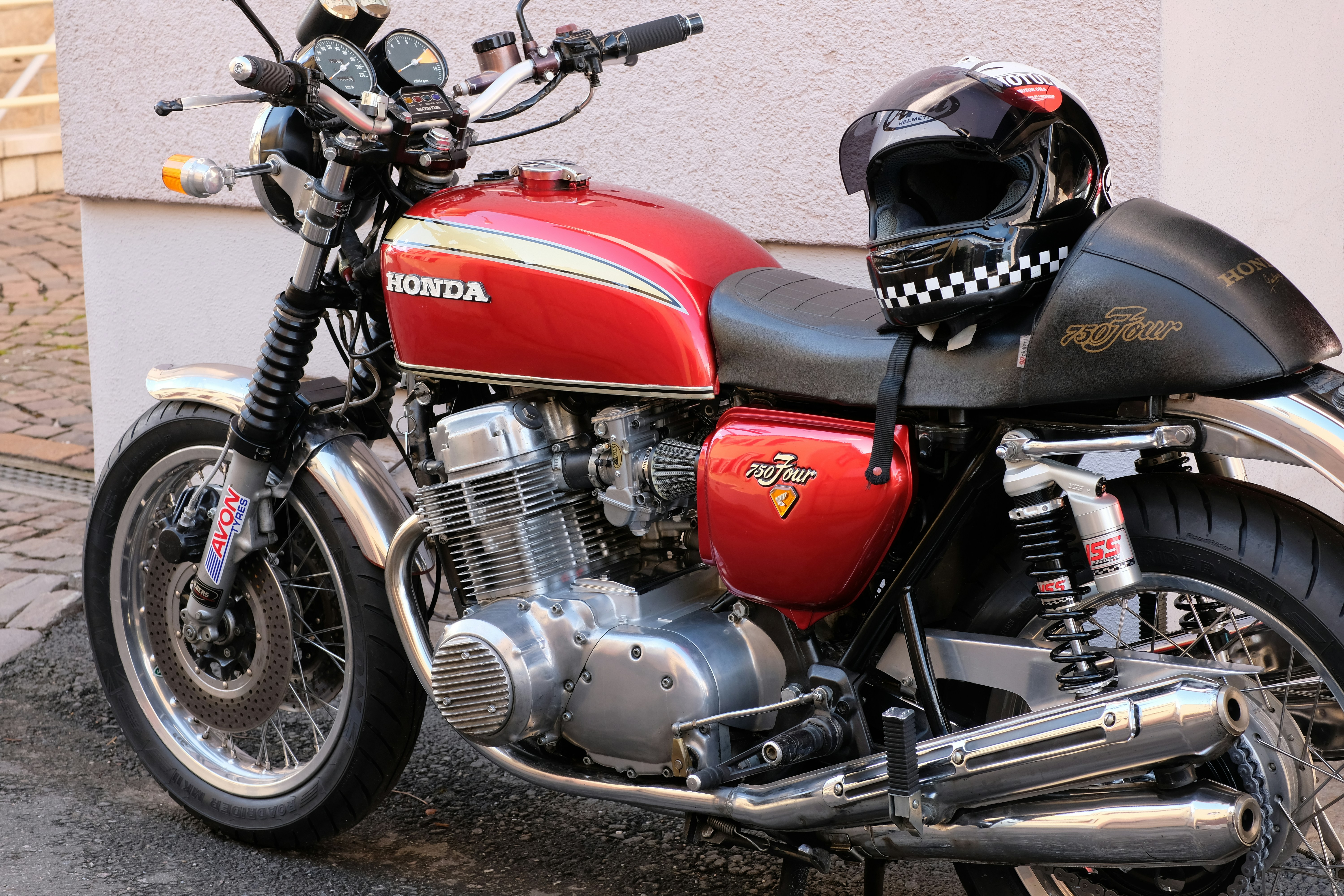 Bright red Honda cafe racer with chrome engine and black-and-white check helmet resting on seat; detailed side view emphasizing the bike's polished metal and vintage styling.