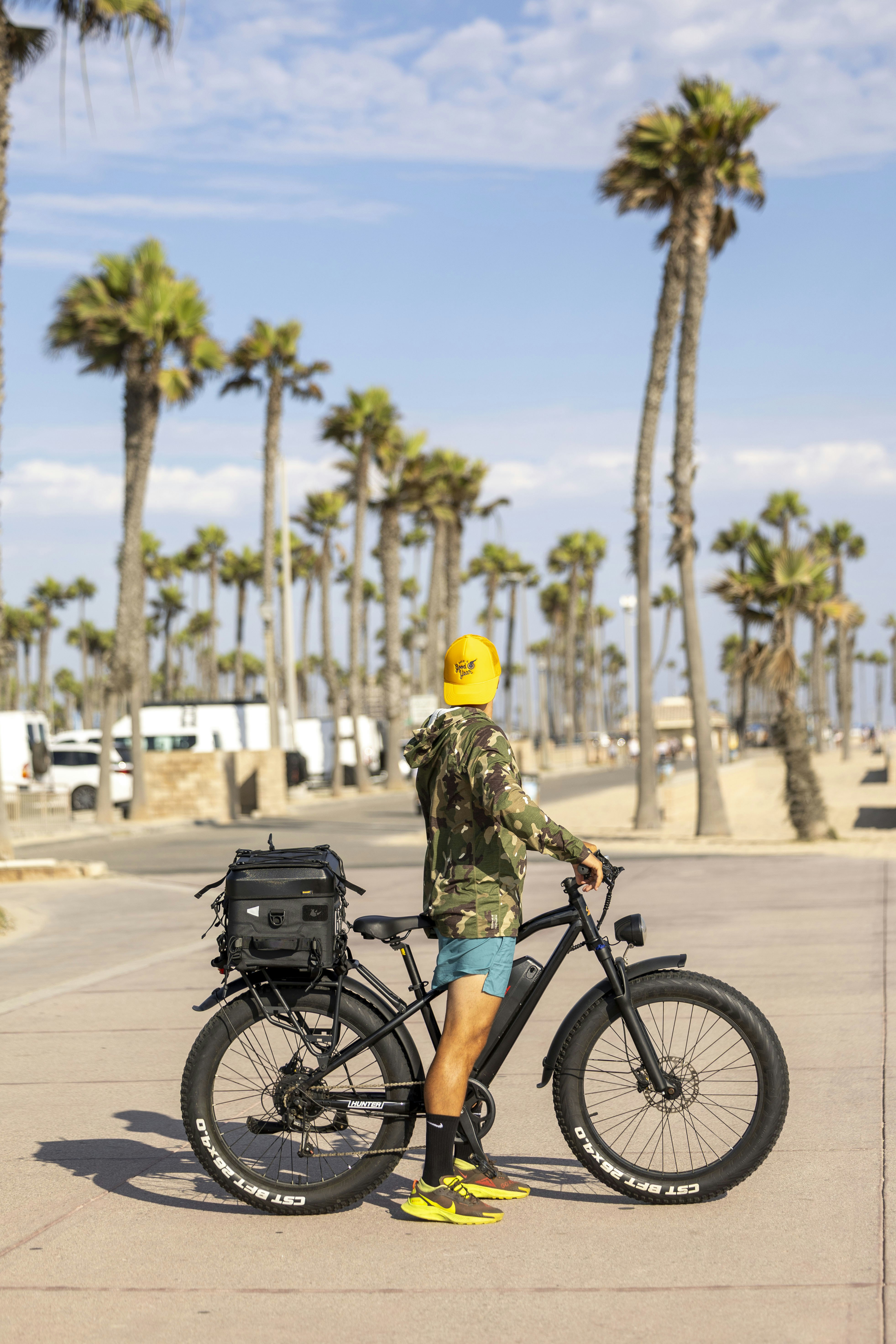 Athletic rider in camo jacket, turquoise shorts, and bright yellow shoes stands beside a fat-tire bike with loaded rear bag under a row of palm trees along a sunny coastal parking lot.