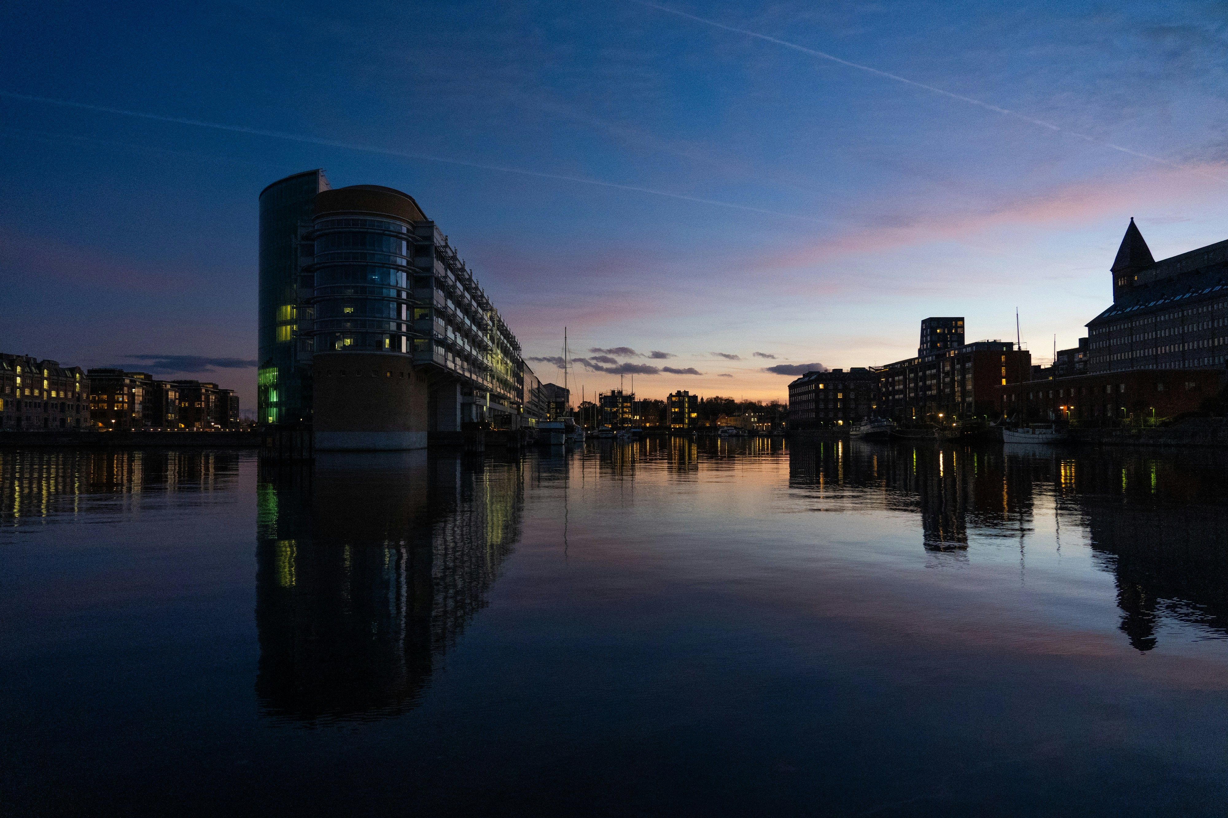 Dusk-lit cityscape reflection across calm river with modern buildings lining the water, capturing the tranquil glow and silhouettes of architecture.