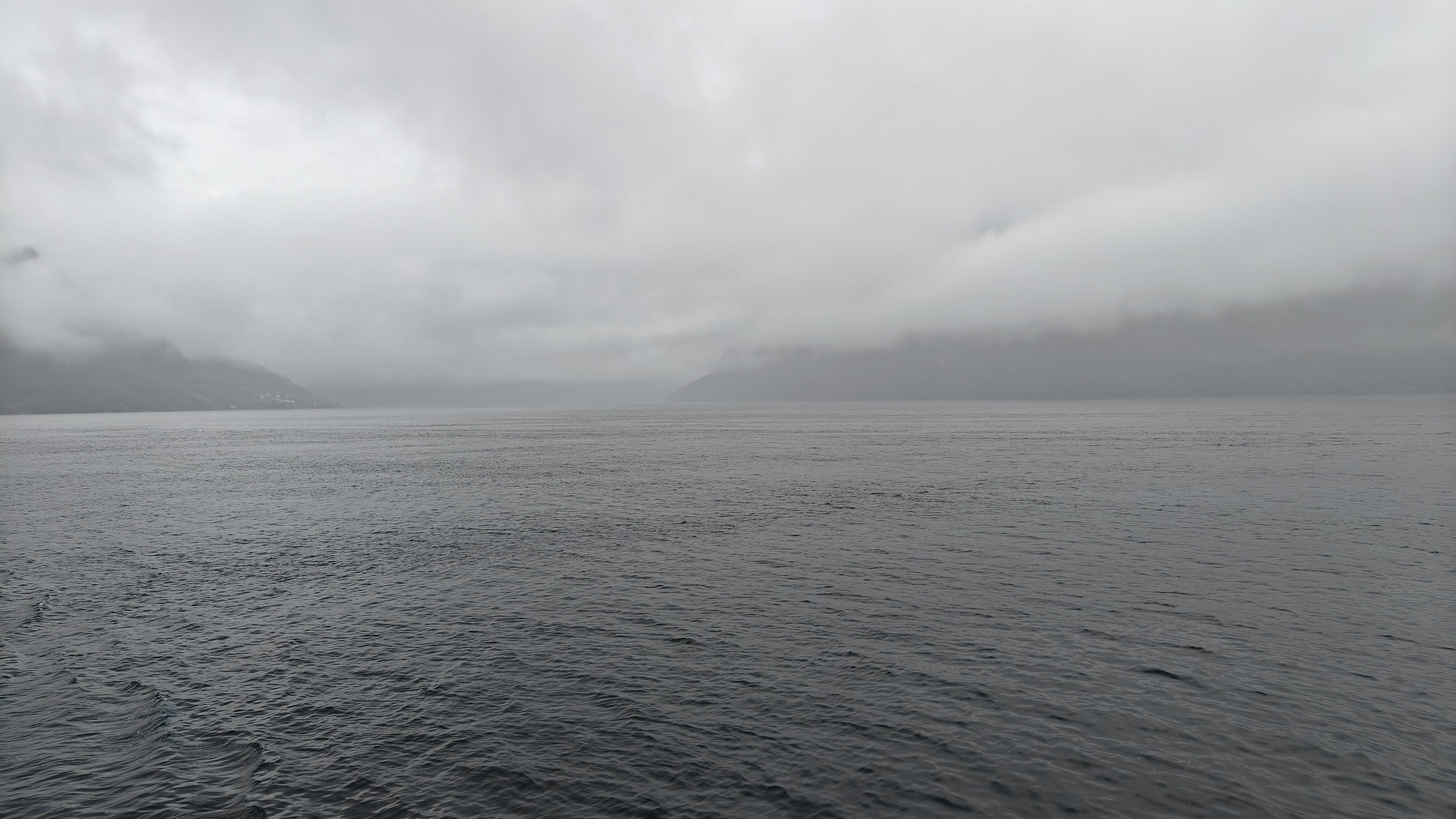 Low-contrast seascape with distant landforms shrouded in fog over a calm body of water.