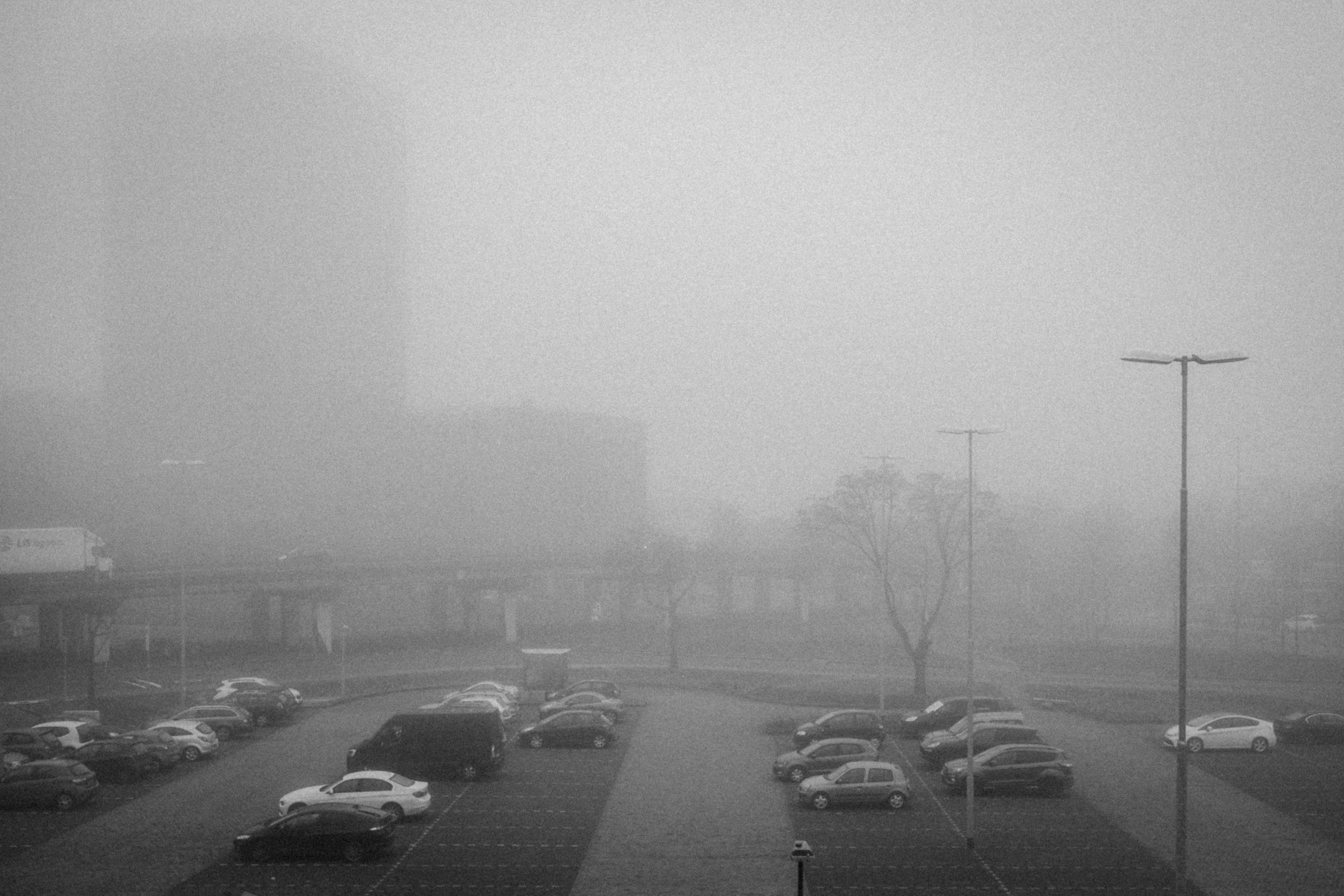 Low-contrast gray-scale scene of a foggy parking lot with scattered cars and a distant, indistinct urban silhouette; the atmosphere emphasizes hush and minimal detail.