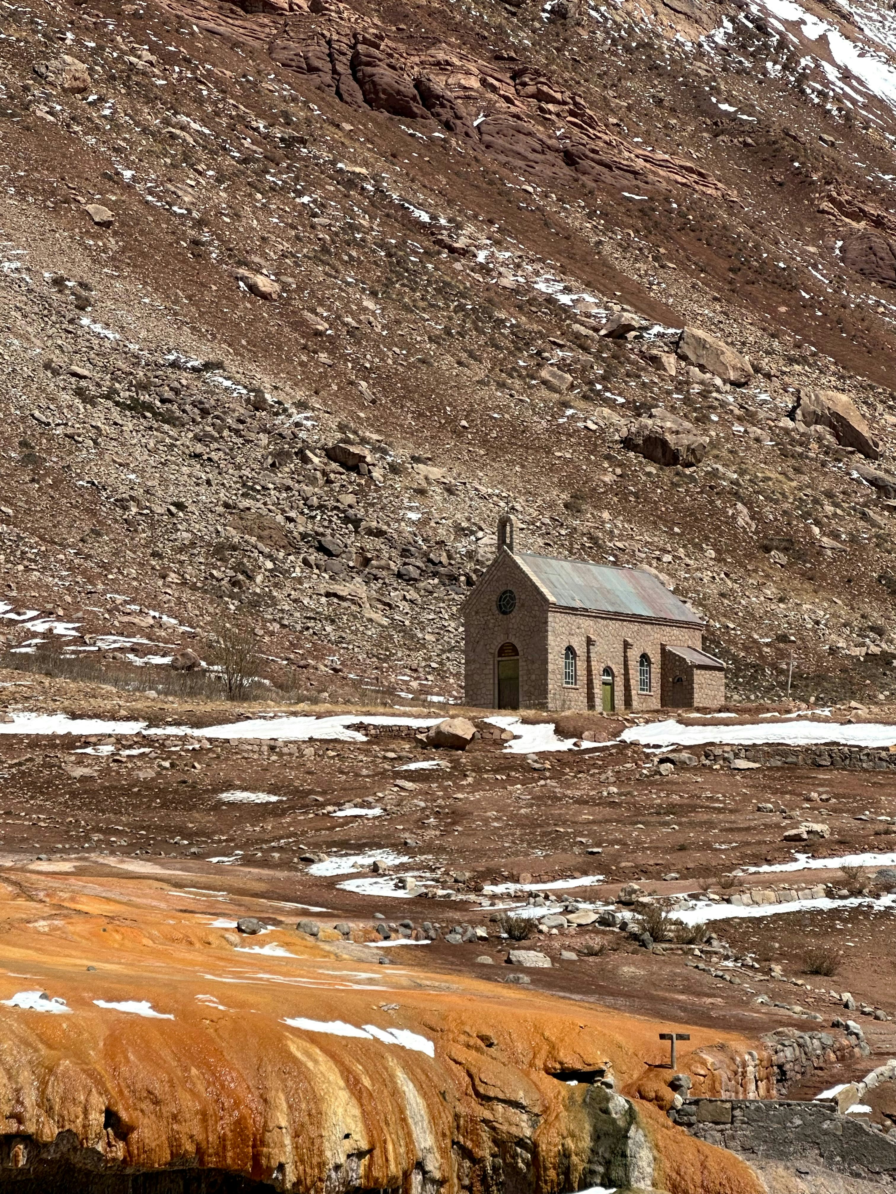 A small stone chapel sits at the base of a rugged, snow-dusted mountainside, with mineral-rich orange and white terraces in the foreground; a solitary, weathered building anchors the remote landscape.