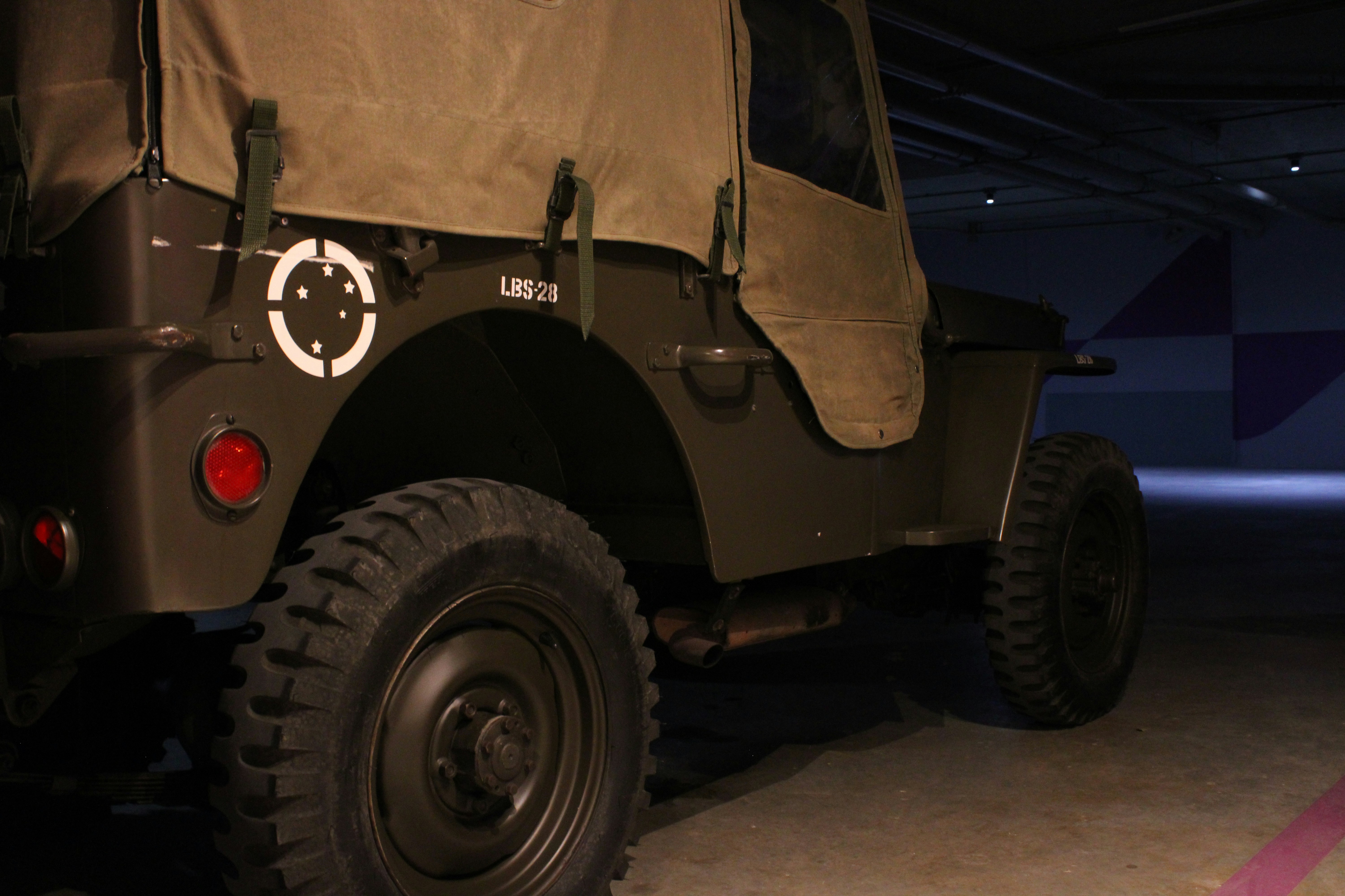 Military jeep under dim hangar lighting with heavy canvas top, rugged tires, and a blue-purple backdrop in the distance.