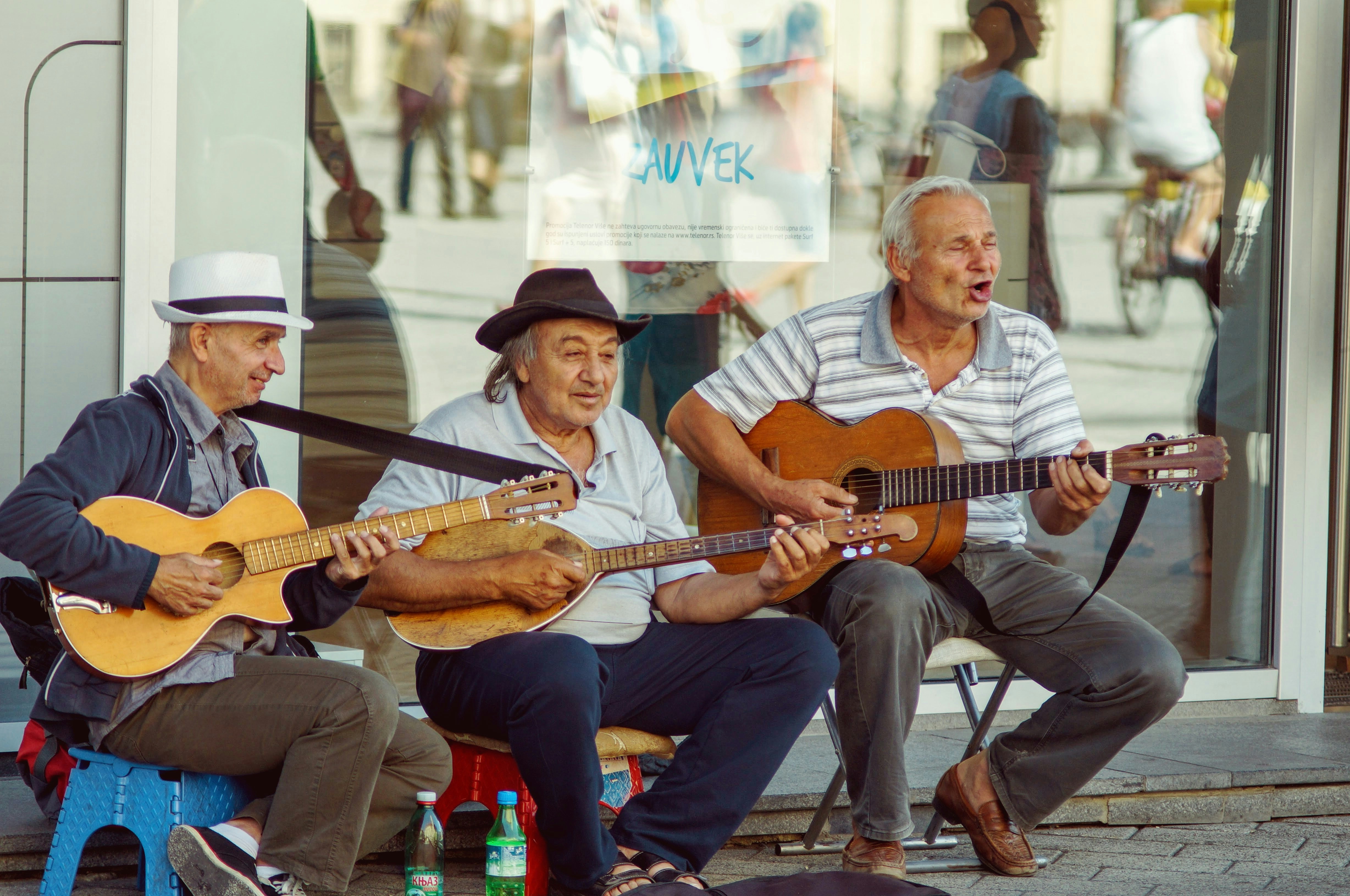Three older men playing acoustic guitars and a tambura on a city sidewalk.