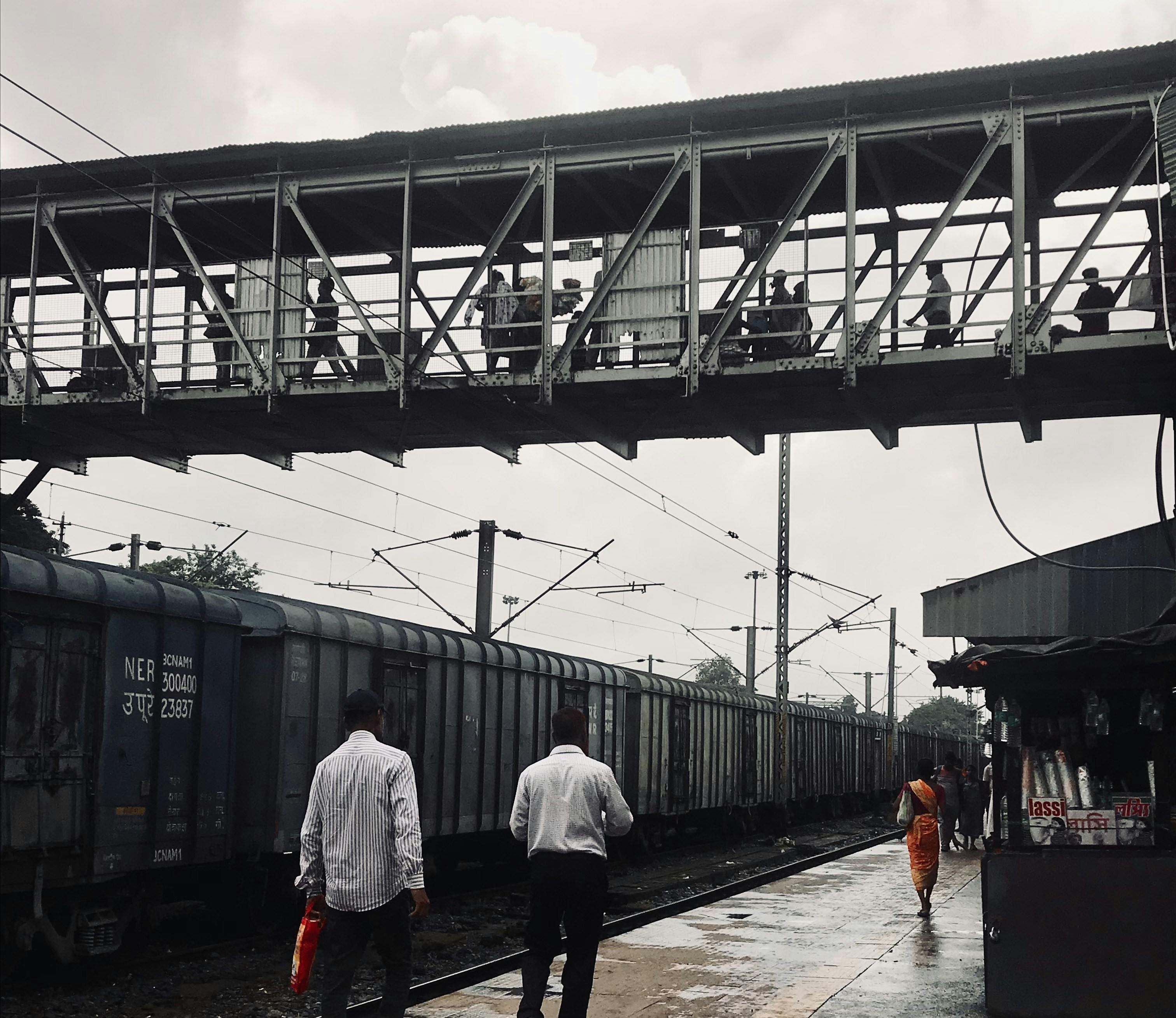 People walking on a metal footbridge above a train at a railway station.