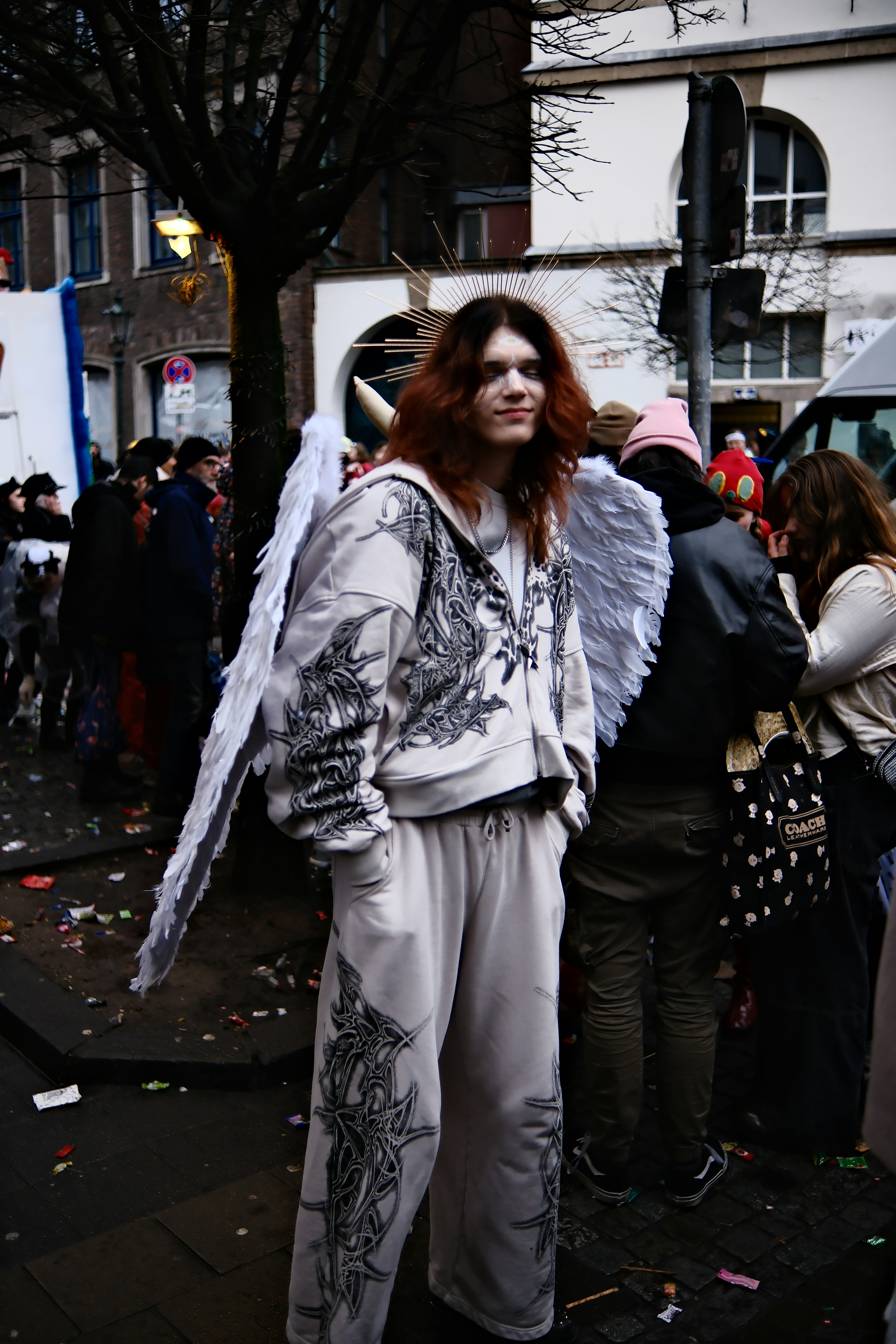 Angel-costumed figure with white feathered wings stands amid a crowded street festival, wearing a gray embroidered tracksuit. The scene captures a lively urban crowd and festive atmosphere.