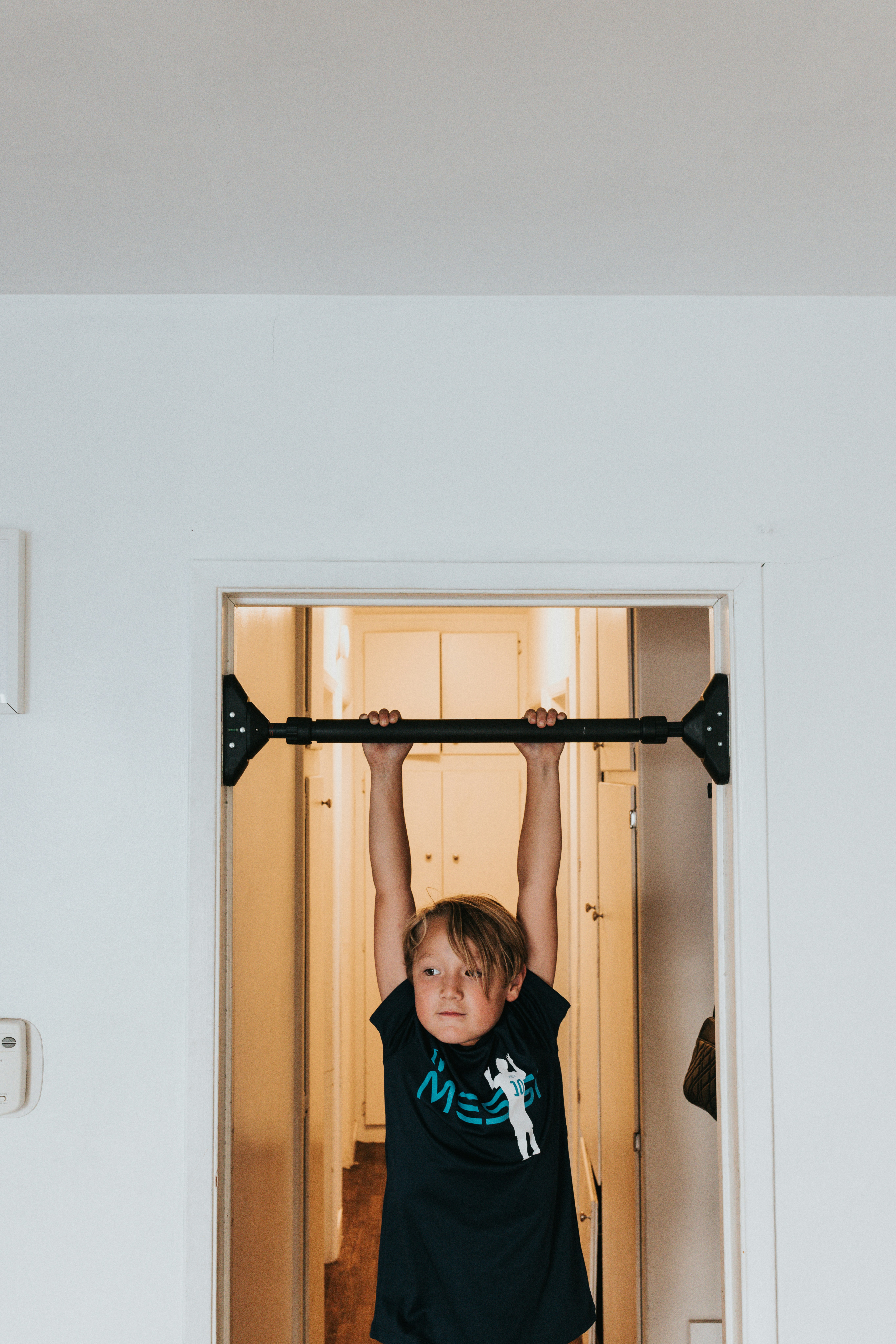 Young boy hanging from a pull-up bar in a doorway, using a narrow indoor corridor as the frame. This is a candid indoor shot capturing effort and concentration.