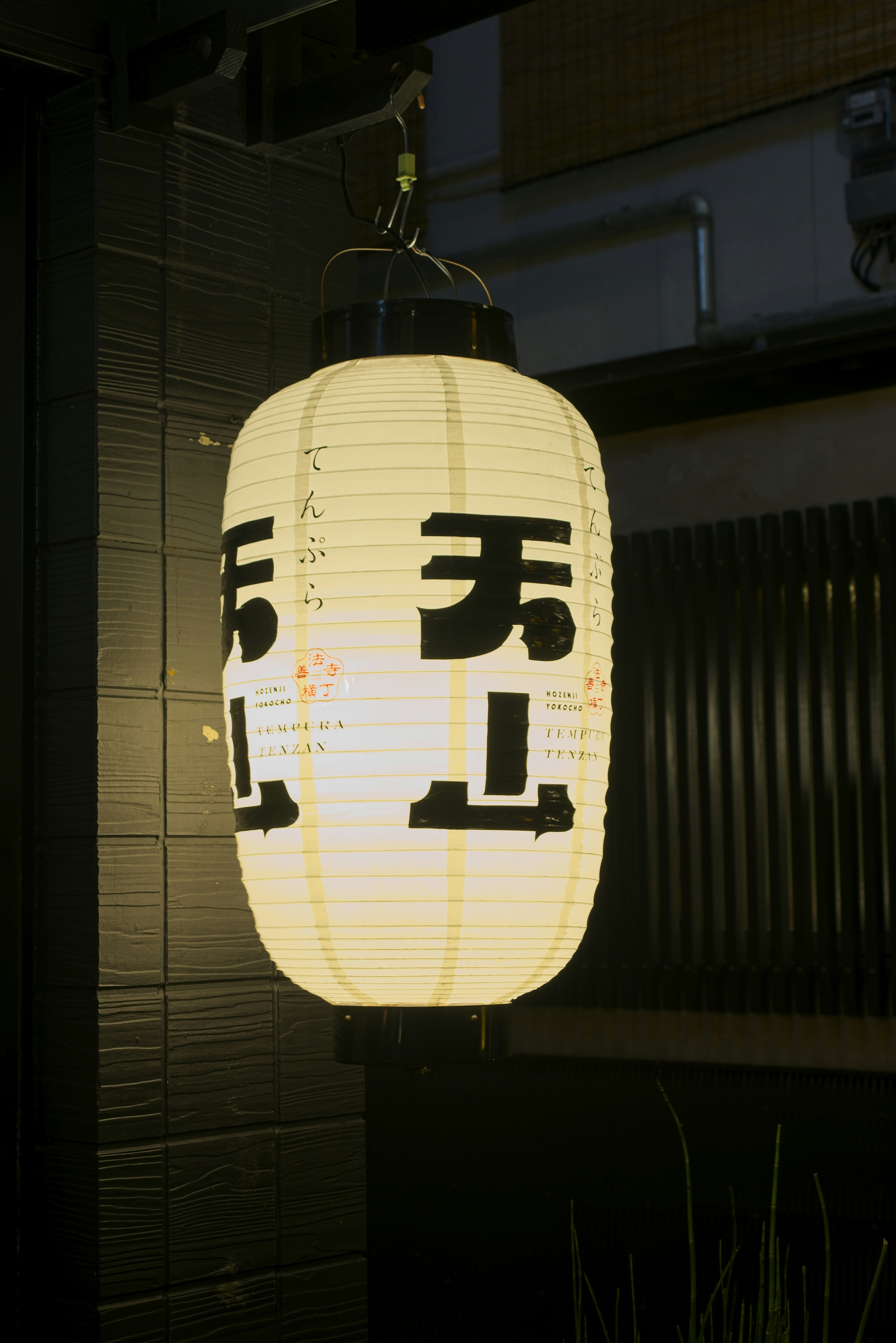 A glowing white paper lantern hangs elegantly outside a tempura restaurant near Dotonbori in Osaka, its bold black kanji reading "Tenzan" illuminated softly against the dark wooden facade on a hot summer evening in July. The warm, minimalist lighting creates a striking contrast with the shadowed background, highlighting the lantern's fine texture and traditional craftsmanship. This quiet, intimate scene reflects the understated elegance often found in Osaka's smaller dining establishments, offering a peaceful contrast to the district's bustling main streets.