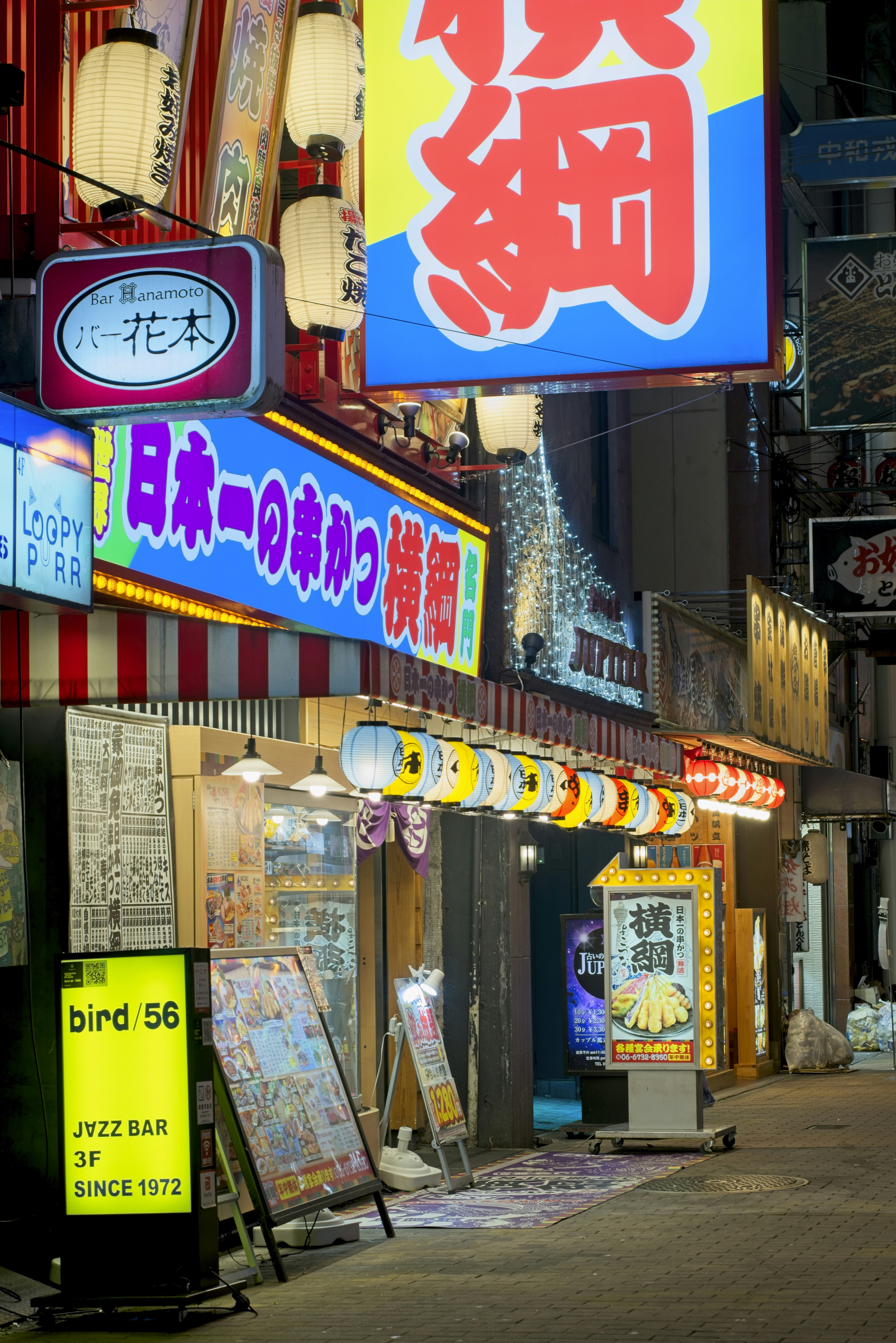 A narrow, atmospheric alley near Dotonbori in Osaka overflows with layered neon signage, colorful lanterns, and vintage advertising on a hot summer evening in July. Bold yellow and red kanji promote kushikatsu restaurants and bars, while a distinctive "bird/56" jazz bar sign since 1972 adds character to the streetscape. Rows of paper lanterns hang beneath a striped awning, leading the eye deeper into the illuminated passageway. The dense, nostalgic mix of signage captures the retro charm and lively energy typical of Osaka's entertainment districts after dark.
