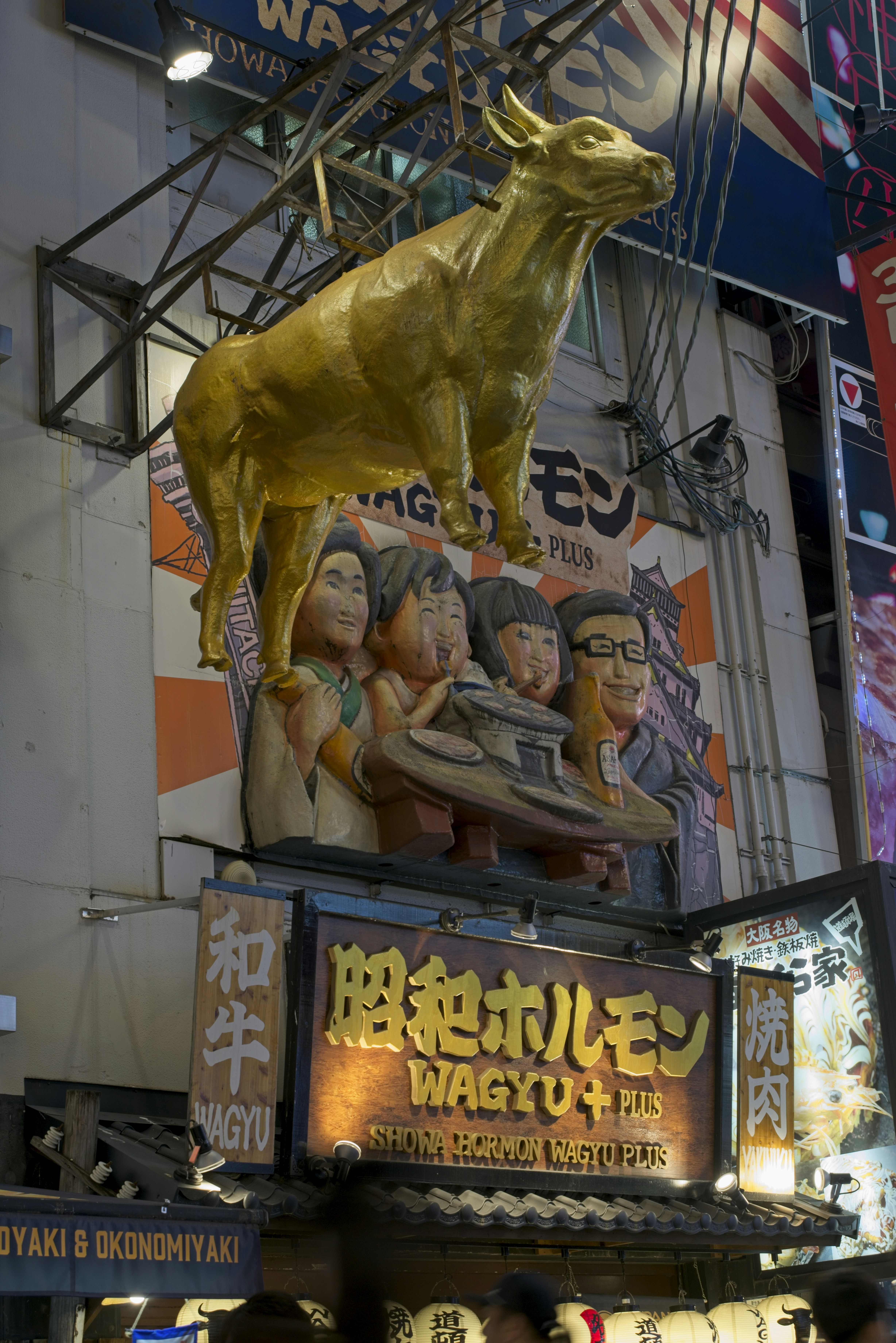 A striking golden bull statue perches above the entrance of Showa Hormon Wagyu Plus near Dotonbori in Osaka, its polished form gleaming against colorful painted characters enjoying a meal below on a hot summer evening in July. Bold neon signage in gold and black advertises the wagyu and yakiniku restaurant, while playful mascots and vibrant graphics decorate the storefront. This eye-catching, larger-than-life display exemplifies the theatrical, over-the-top signage style that makes Osaka's dining district so visually distinctive after dark.