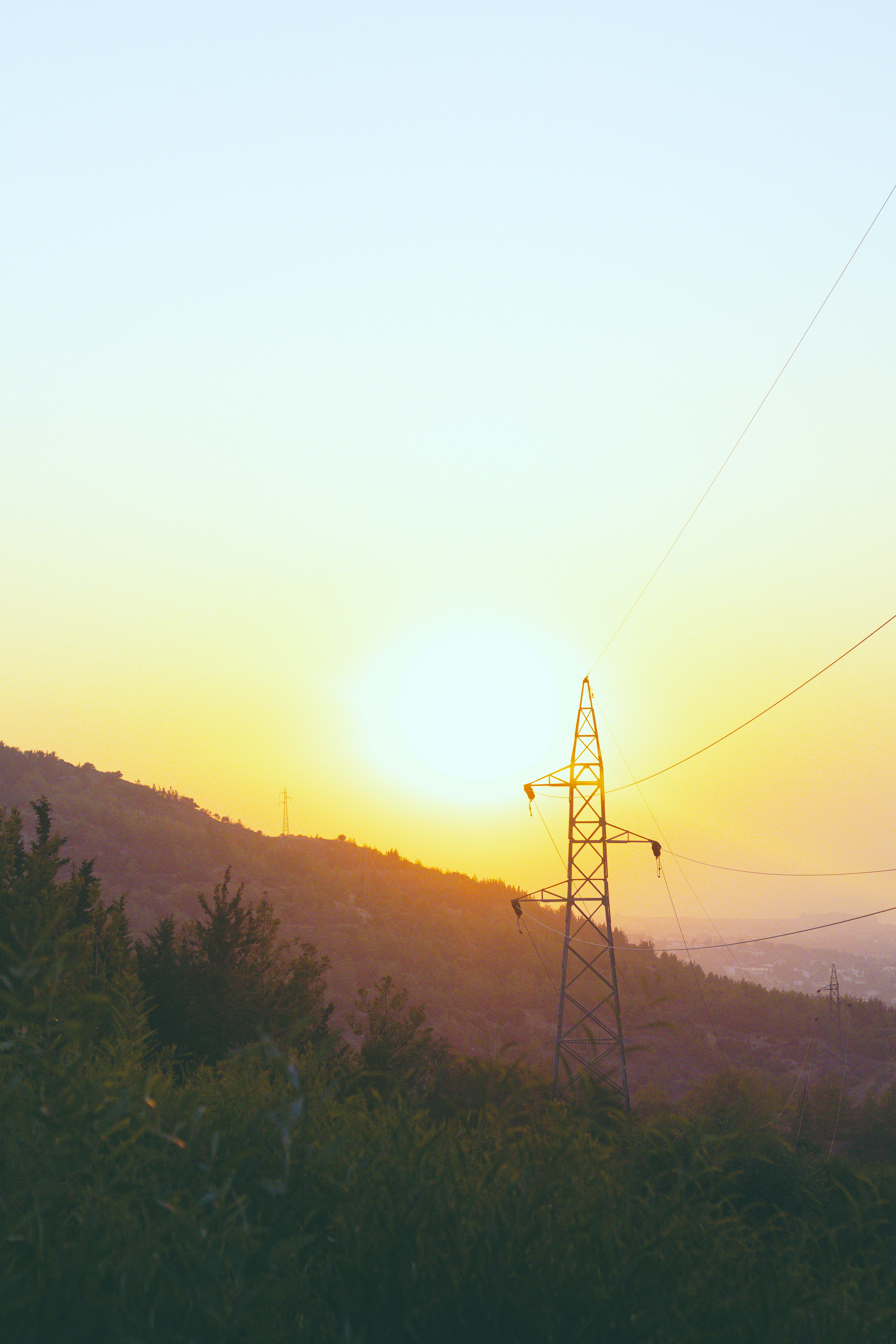 Golden-hour scene featuring a transmission tower and power lines cutting across a hillside under a bright sky. A serene landscape with warm hues and a layered sky, suggesting late-day activity.