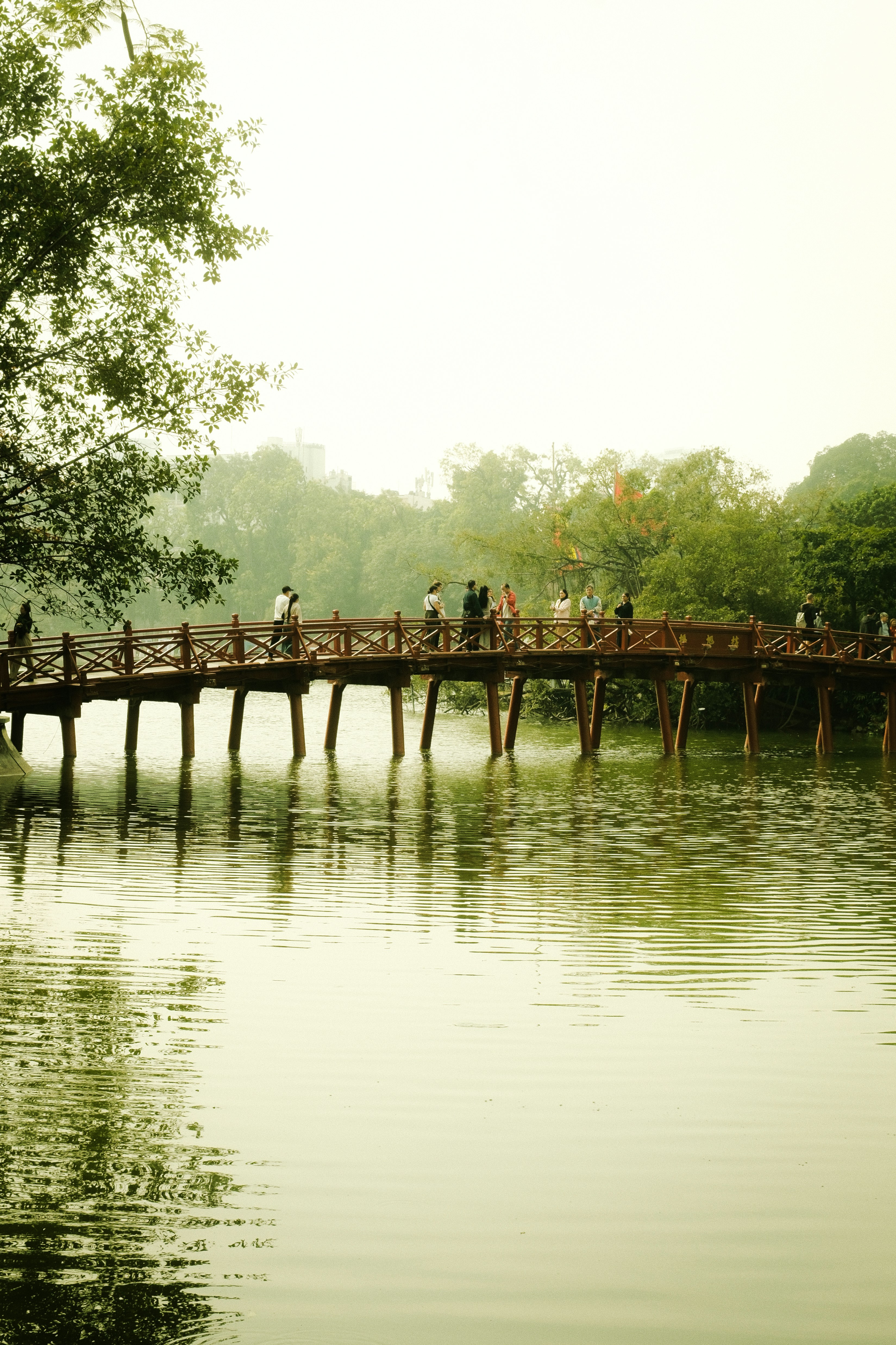 A long wooden footbridge arches over calm water, with guests strolling along its rails as trees frame the misty park in the background.