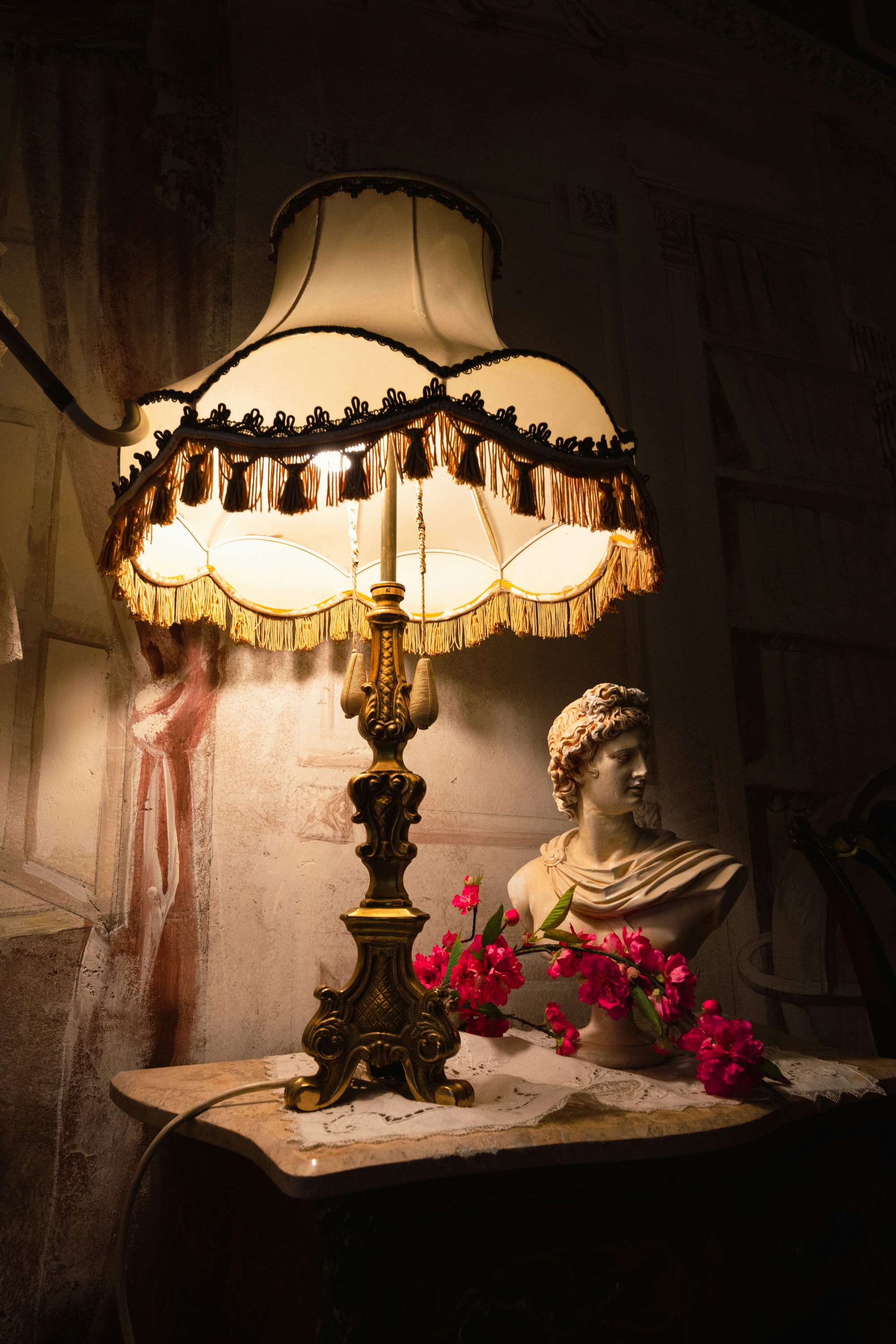 An ornate table lamp with a fringed shade beside a classical bust and flowers.
