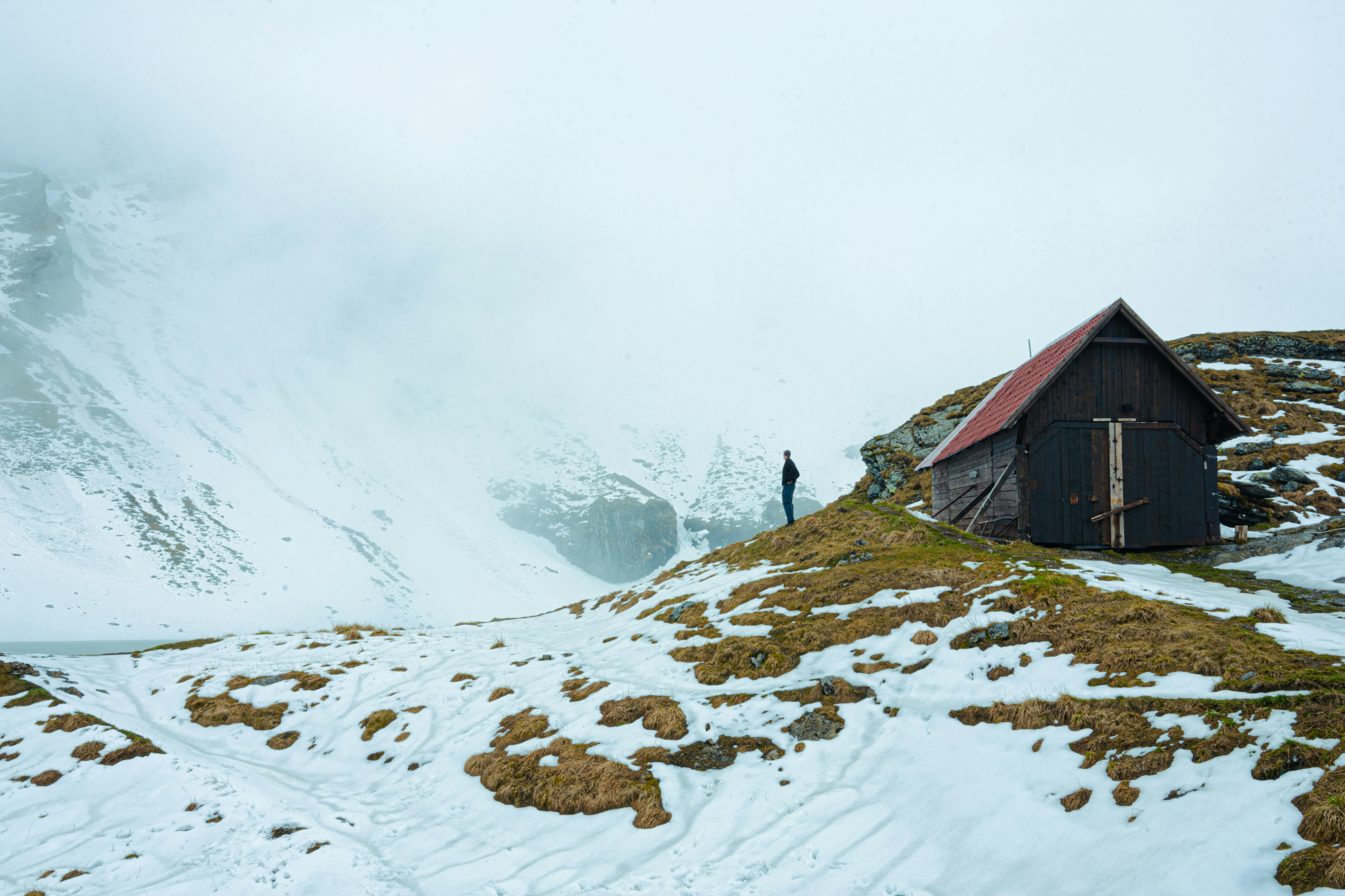  lone figure on a windswept ledge beside a dark cabin in a wintery alpine landscape
