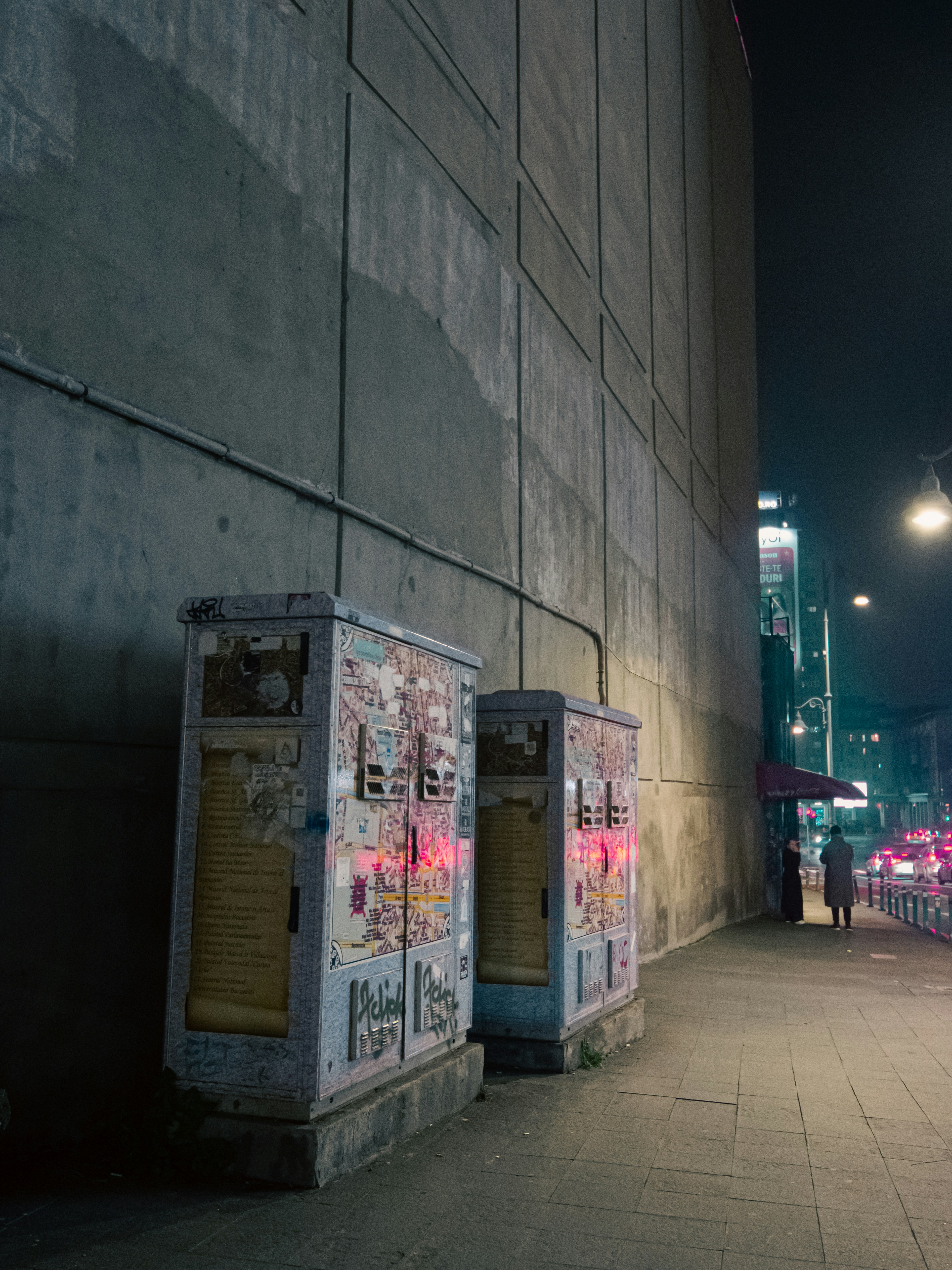 Night street scene with two graffiti-covered kiosks along a tall concrete wall; distant pedestrians and neon light reflections hint at urban life after dark.