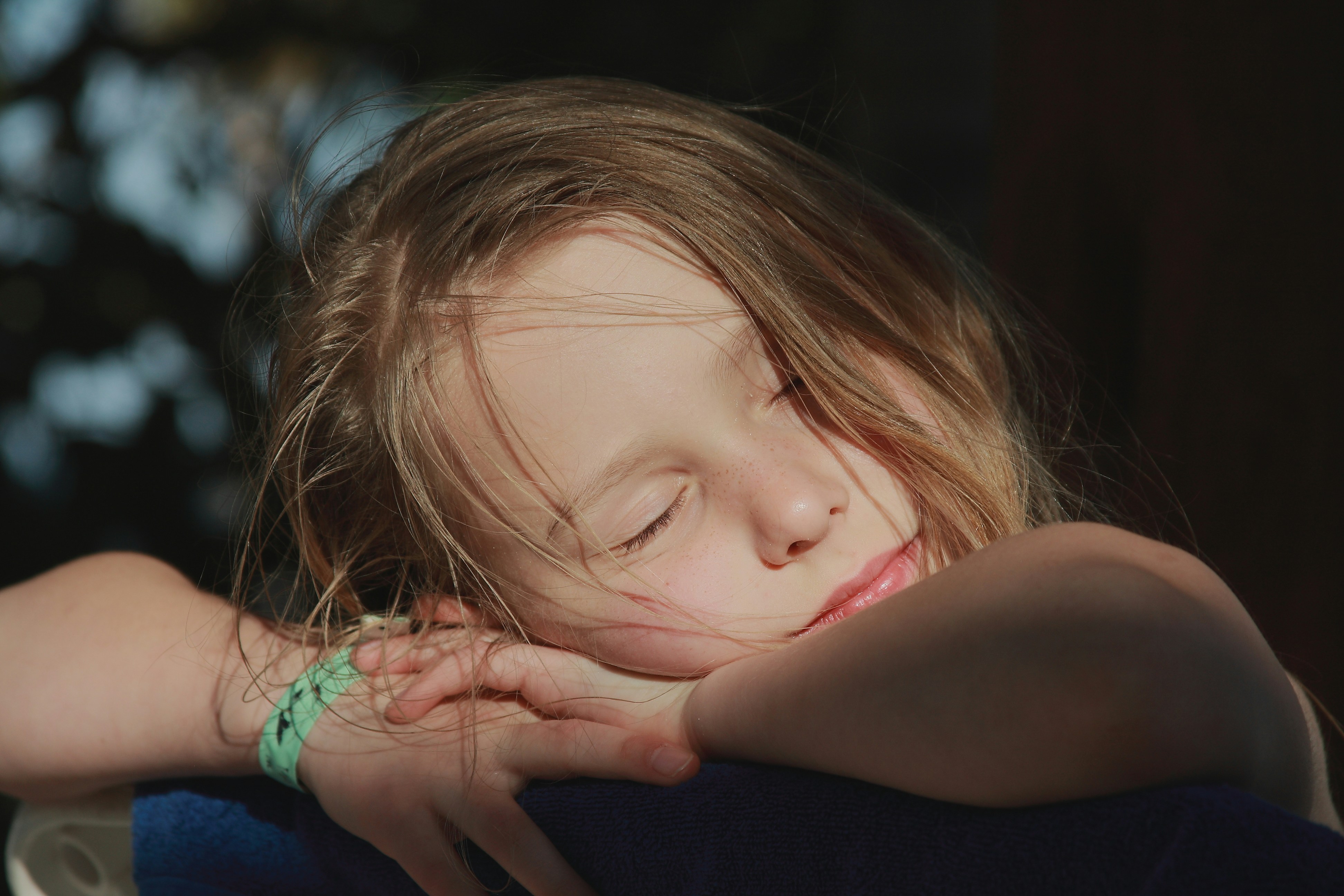 Young girl rests with eyes closed, head tilted on folded arms; warm sunlight and shadow create a gentle, intimate portrait. Photograph type: candid portrait.