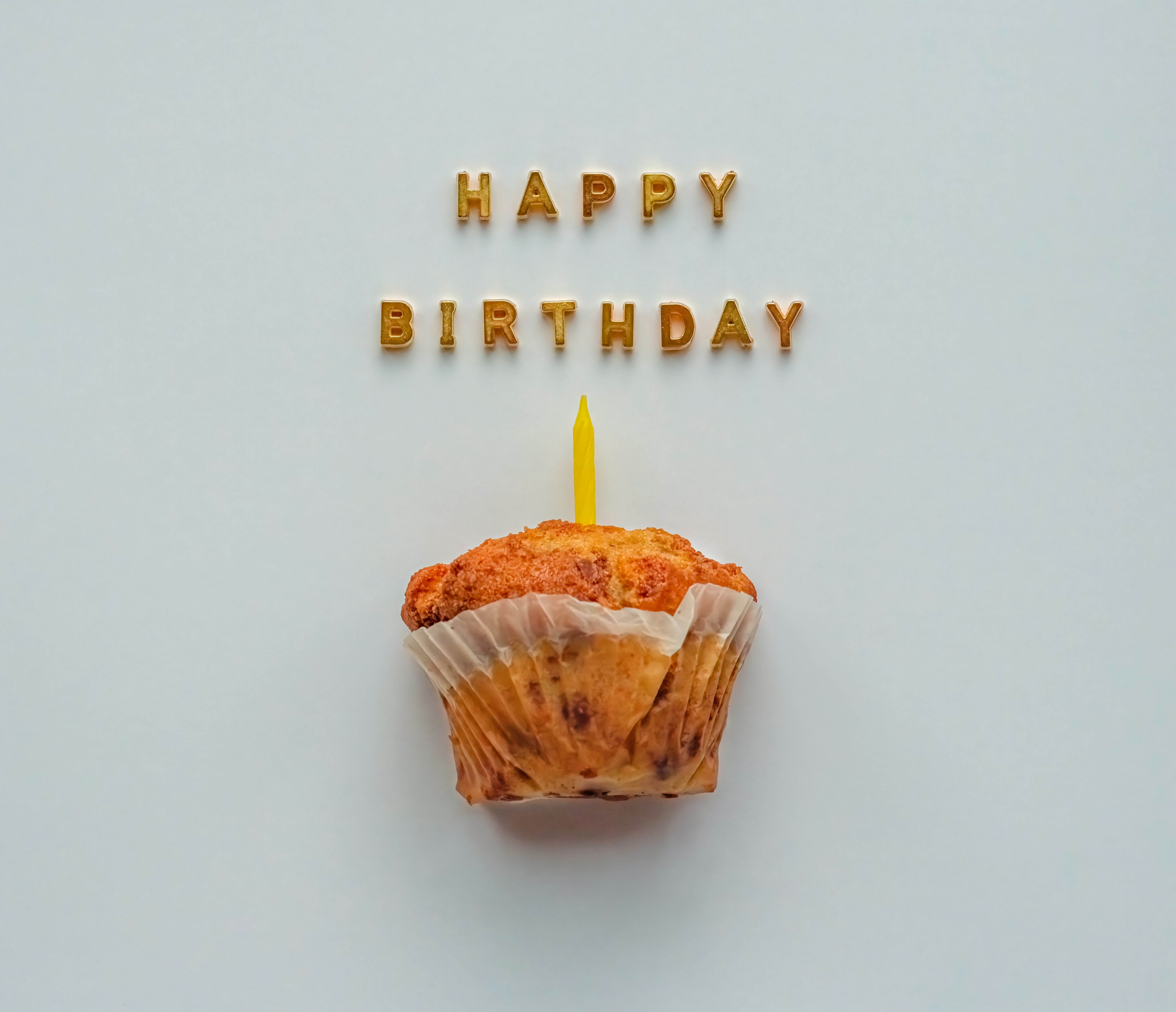A muffin with a single yellow candle below gold letters spelling Happy Birthday.