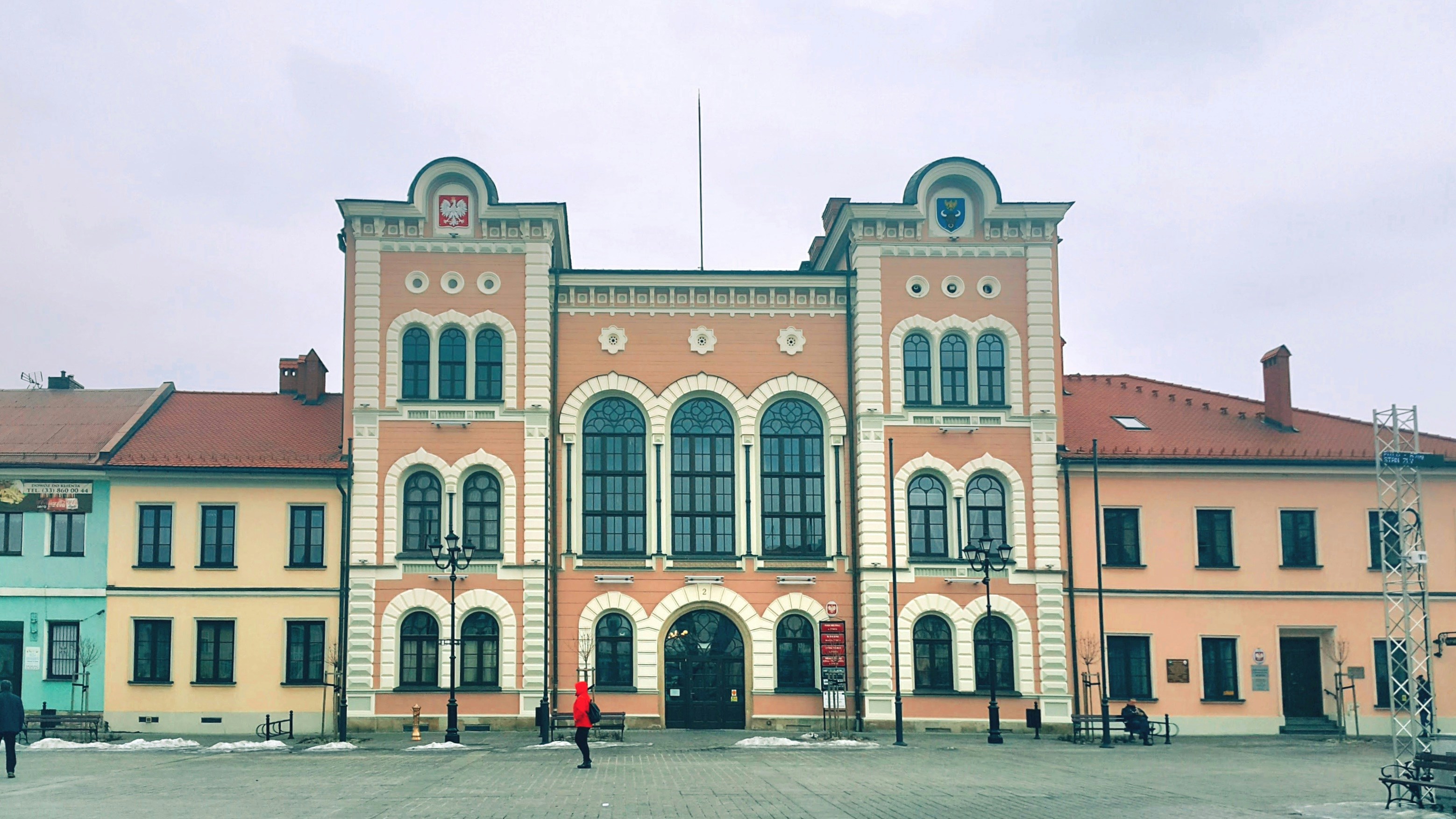 Brick-and-arched town hall façade with pale trim and tall central windows, flanked by pastel adjacent structures; a lone pedestrian at the foreground adds scale.