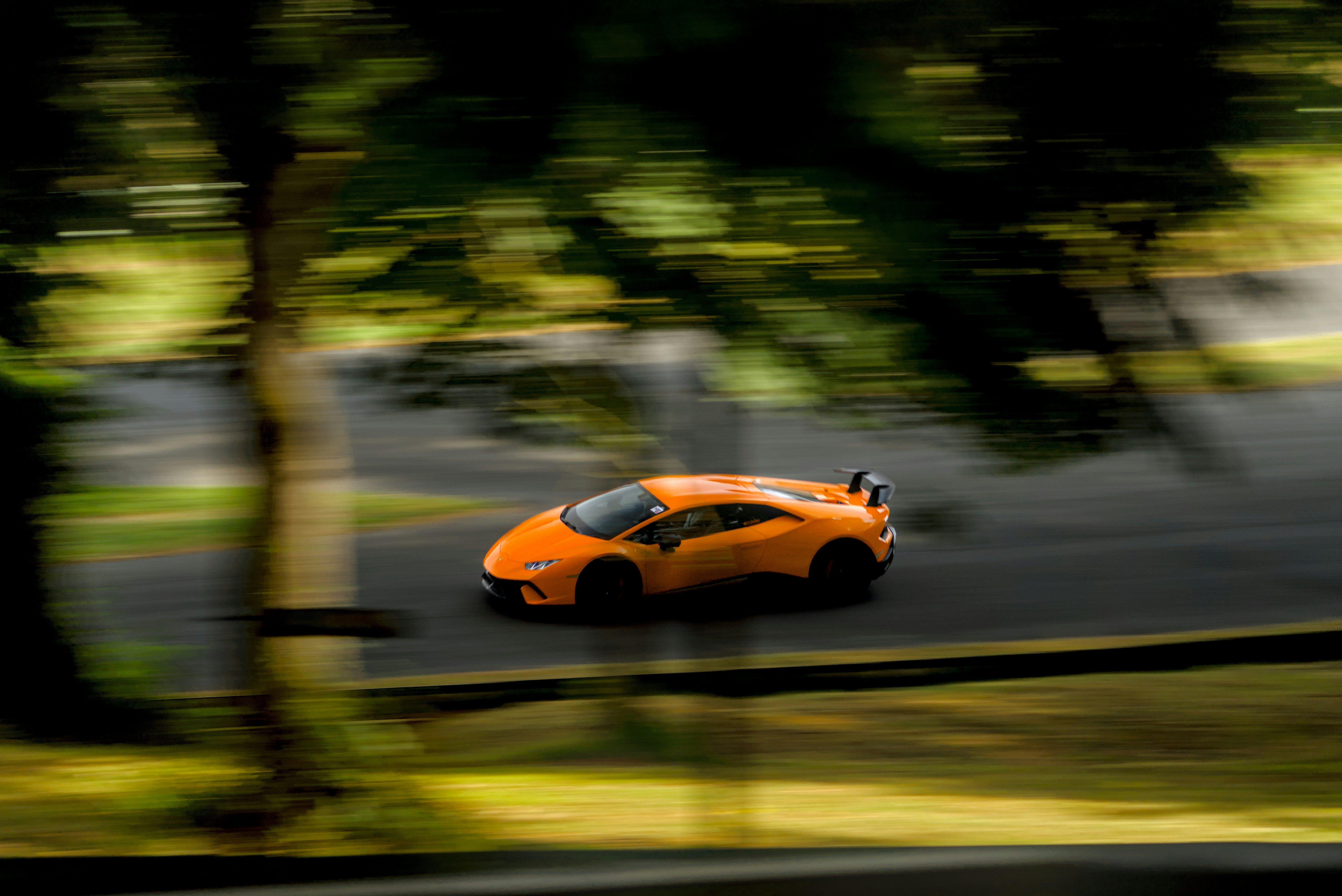 Bright orange sports car speeding along a road with motion blur and surrounding greenery; the vehicle is the focal element in the scene and the emphasis is on speed and motion.