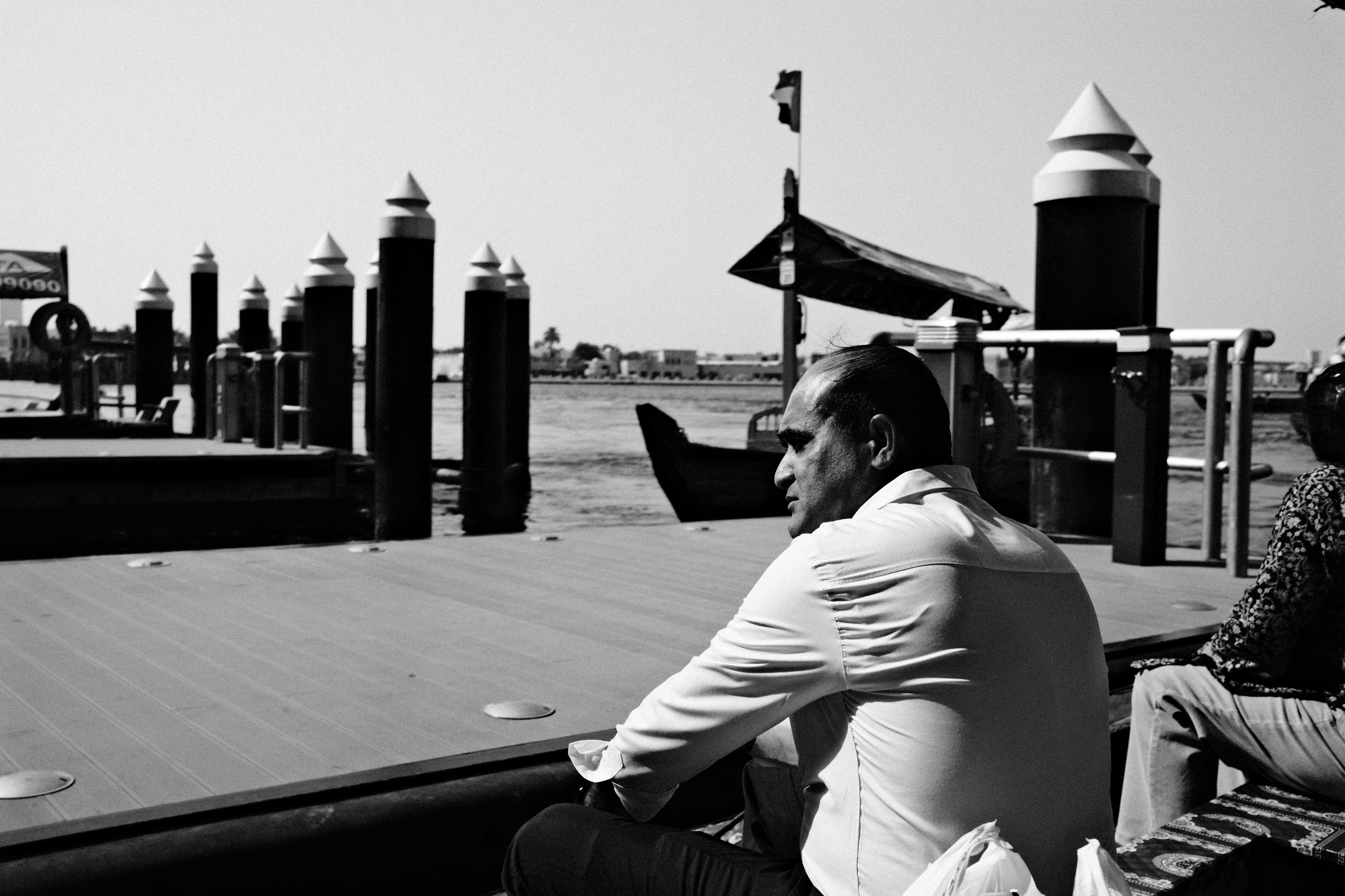 A man sits on a dockside bench, facing away, with boats and pilings in the background; the scene is captured in monochrome to emphasize texture and mood. Note: photograph appears as a candid street/harbor scene.