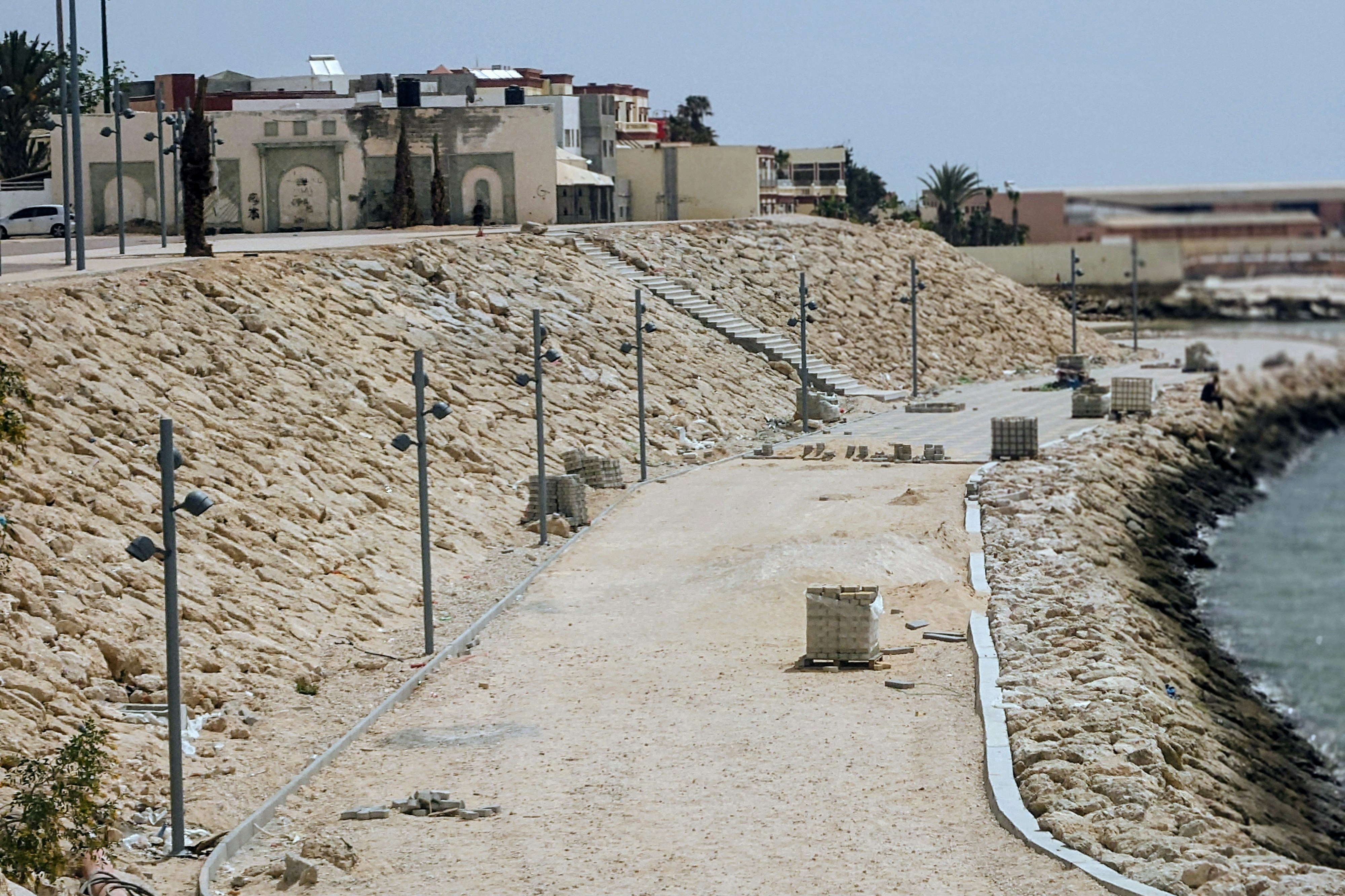 Sunlit stone seawall path along the shoreline with lamp posts and distant buildings; calm sea to the right and a clear sky above. A quiet coastal walkway emphasizing the built edge against the water.