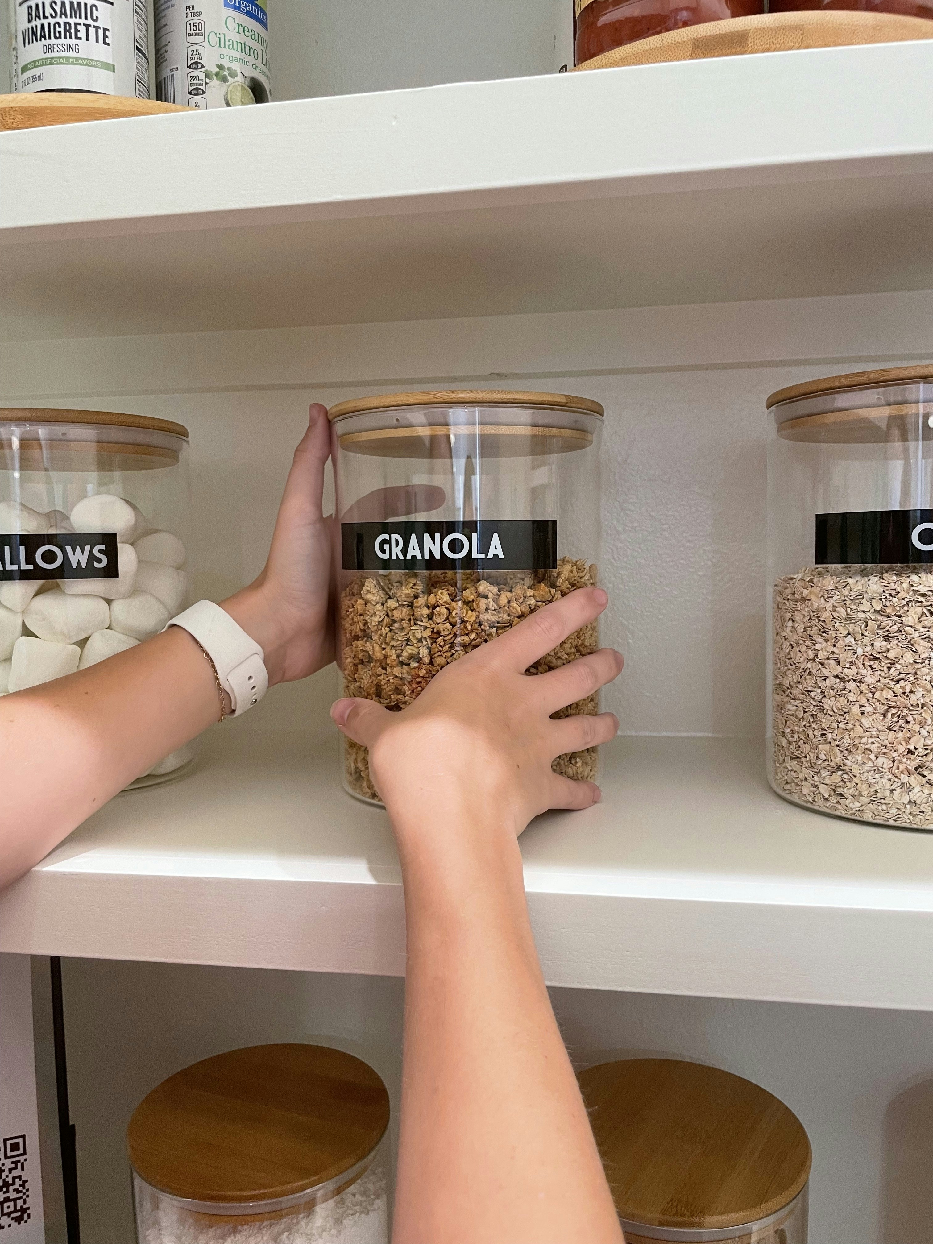 Hands adjust a glass canister labeled GRANOLA on a shelf in a tidy pantry; soft, warm lighting highlights the scene with other labeled jars in view.