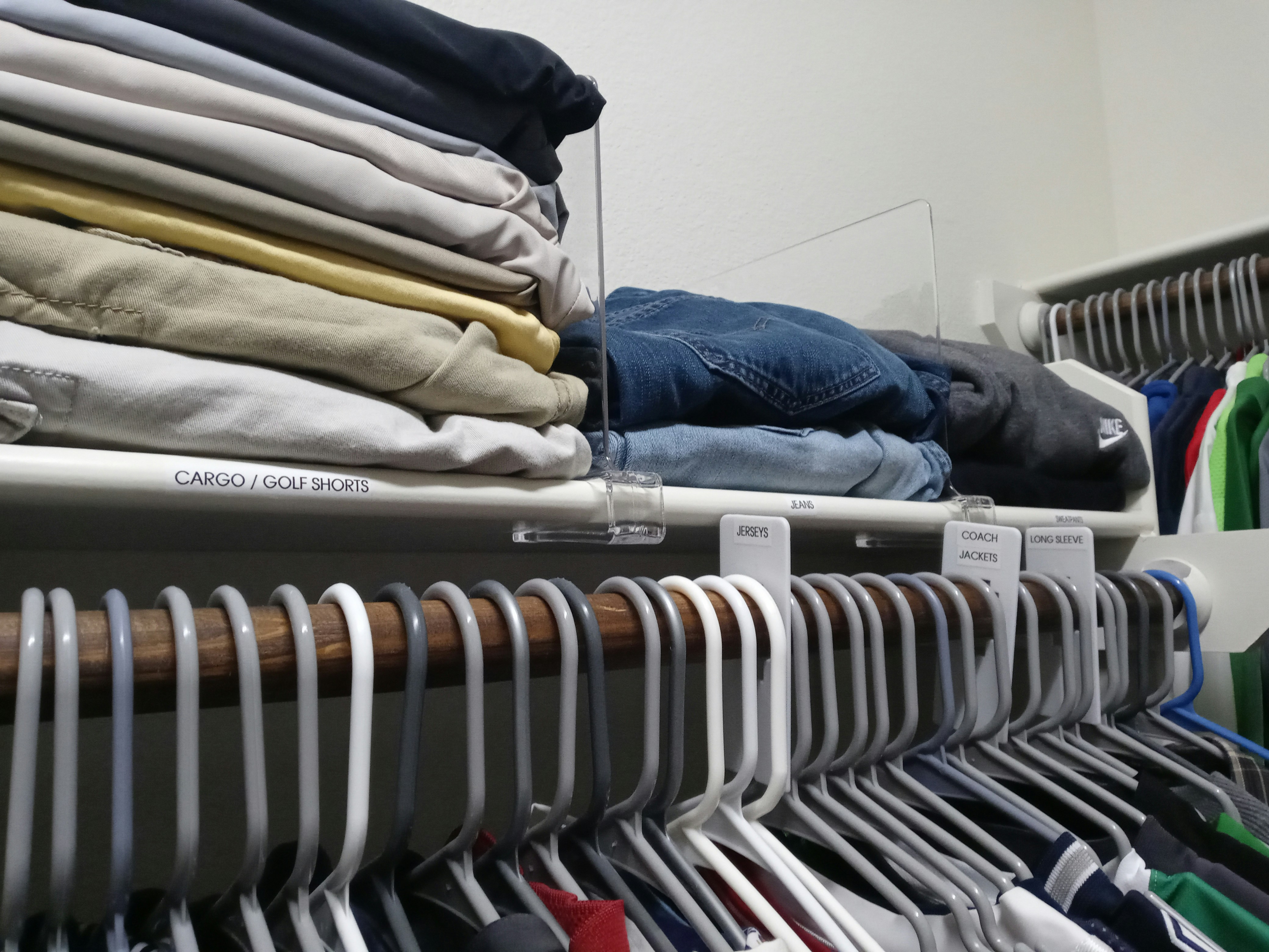 Shelf of folded clothes above a row of hangers in a tidy closet, showing labels and a mix of garments.