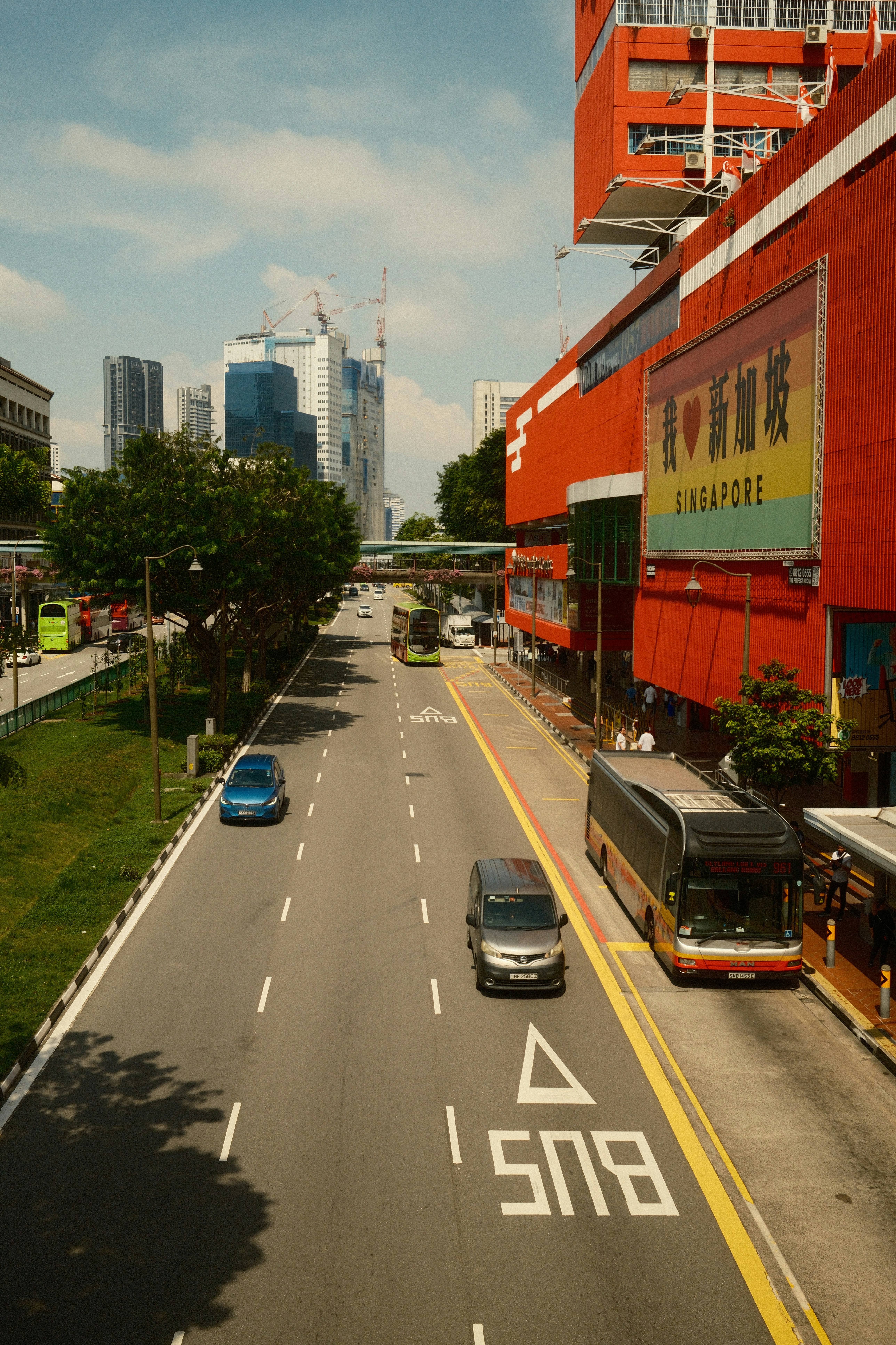 Bus-stop street scene with a bold red building on the right, a multi-lane road, and greenery along the left. A clear sky and distant skyscrapers hint at a busy urban center.