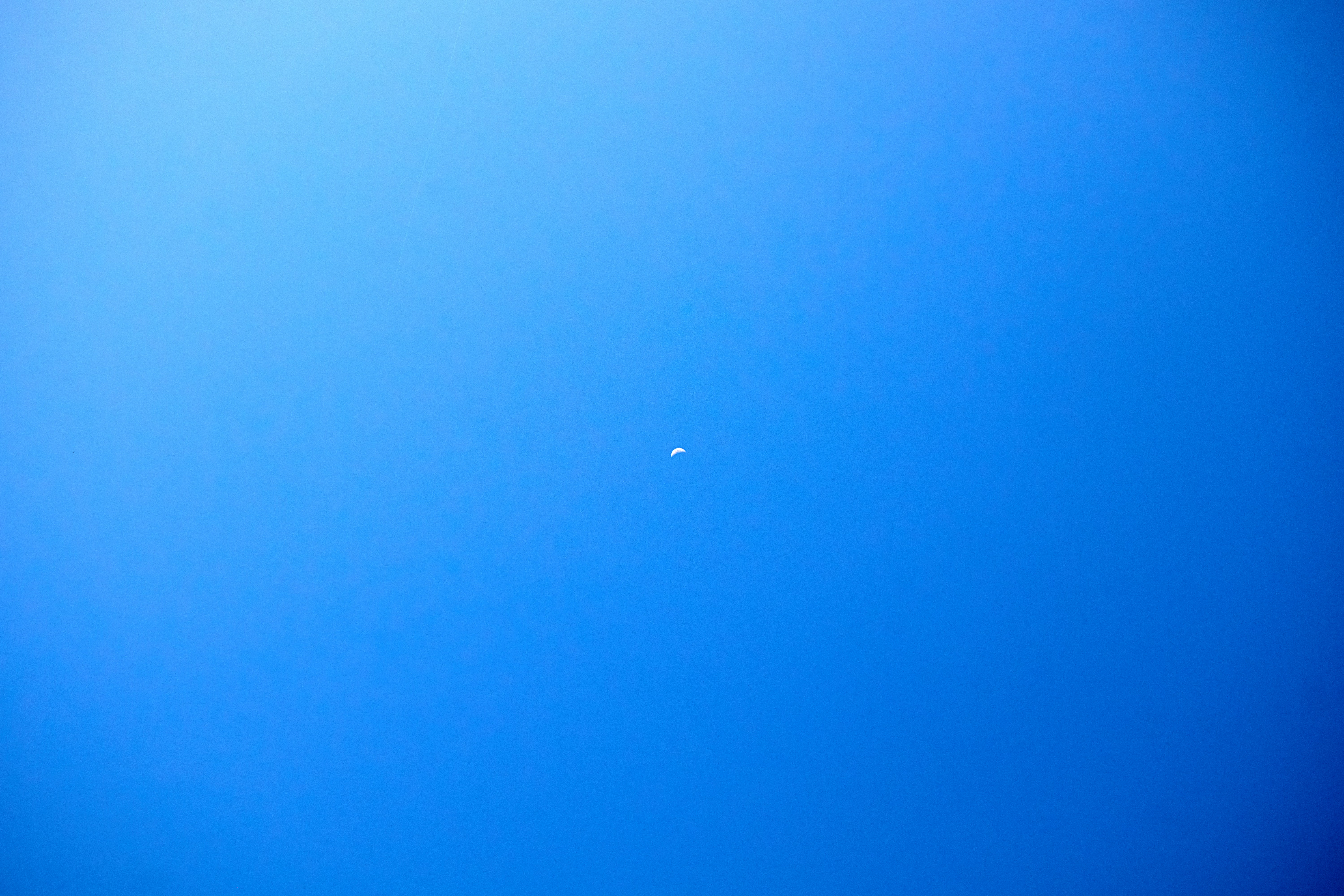 A tiny crescent moon visible in a vast, clear blue sky.
