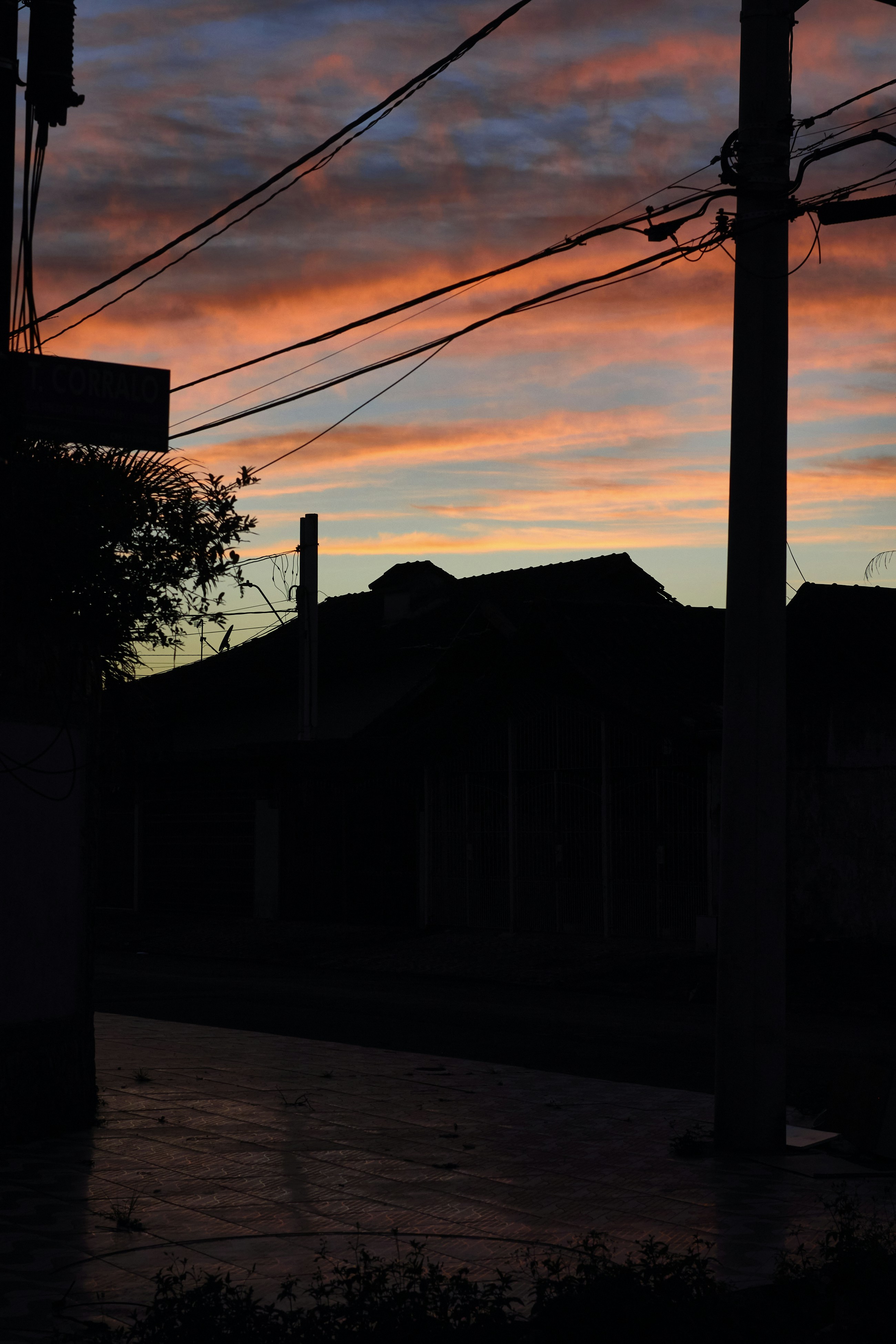 Silhouetted rooftops and power lines against a vibrant orange-pink sunset sky, creating a moody urban silhouette. The scene emphasizes horizon tension and architectural shapes in low light.