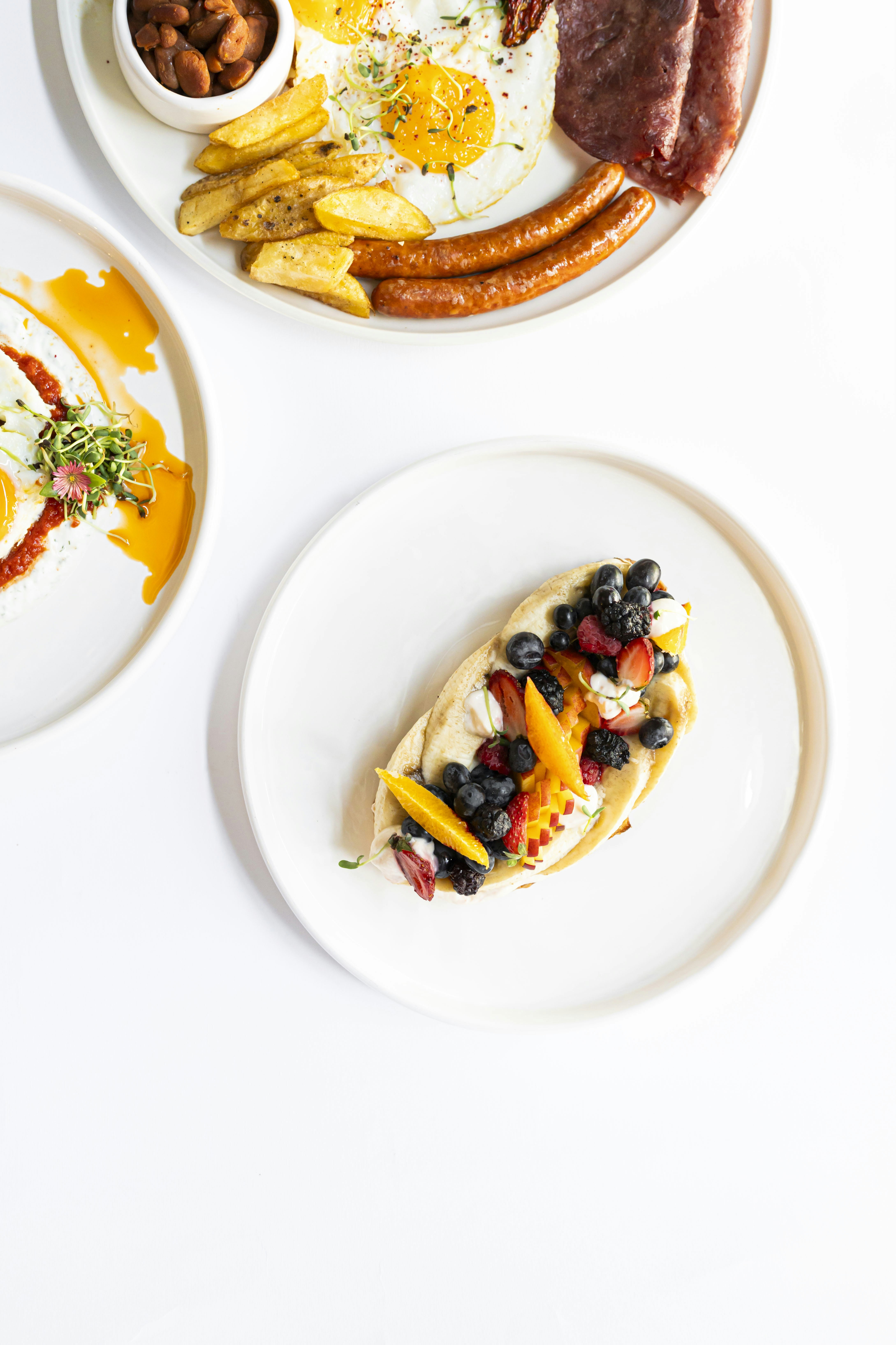 Three plated dishes arranged diagonally with eggs and sides on top left, a stack of pancakes with fruit on a white plate bottom right, and a bright, clean background.