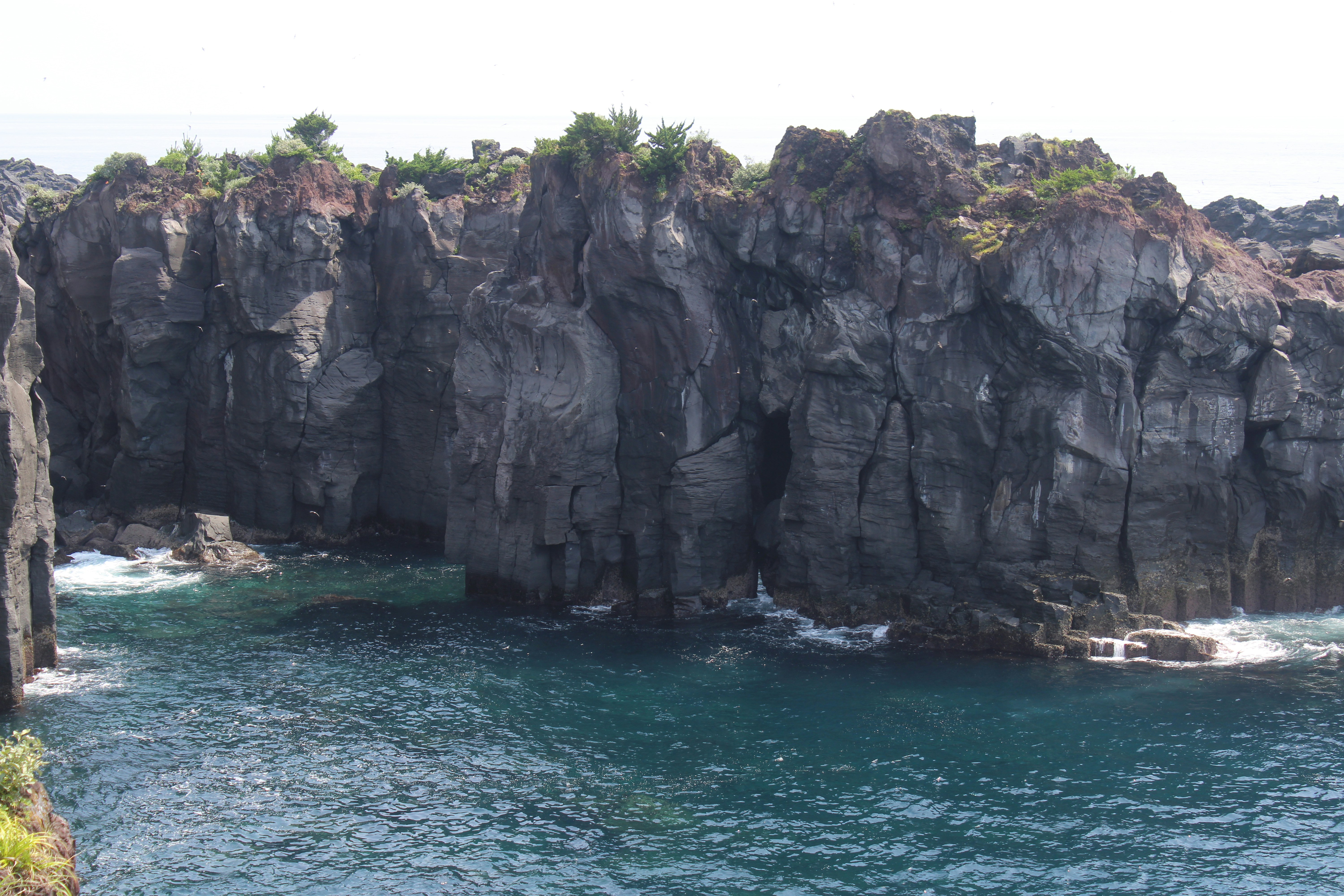 Jagged basalt cliffs rise from clear coastal water, with green vegetation along the rim and a narrow channel at the base. A rugged shoreline and blue-green sea define the scene.
