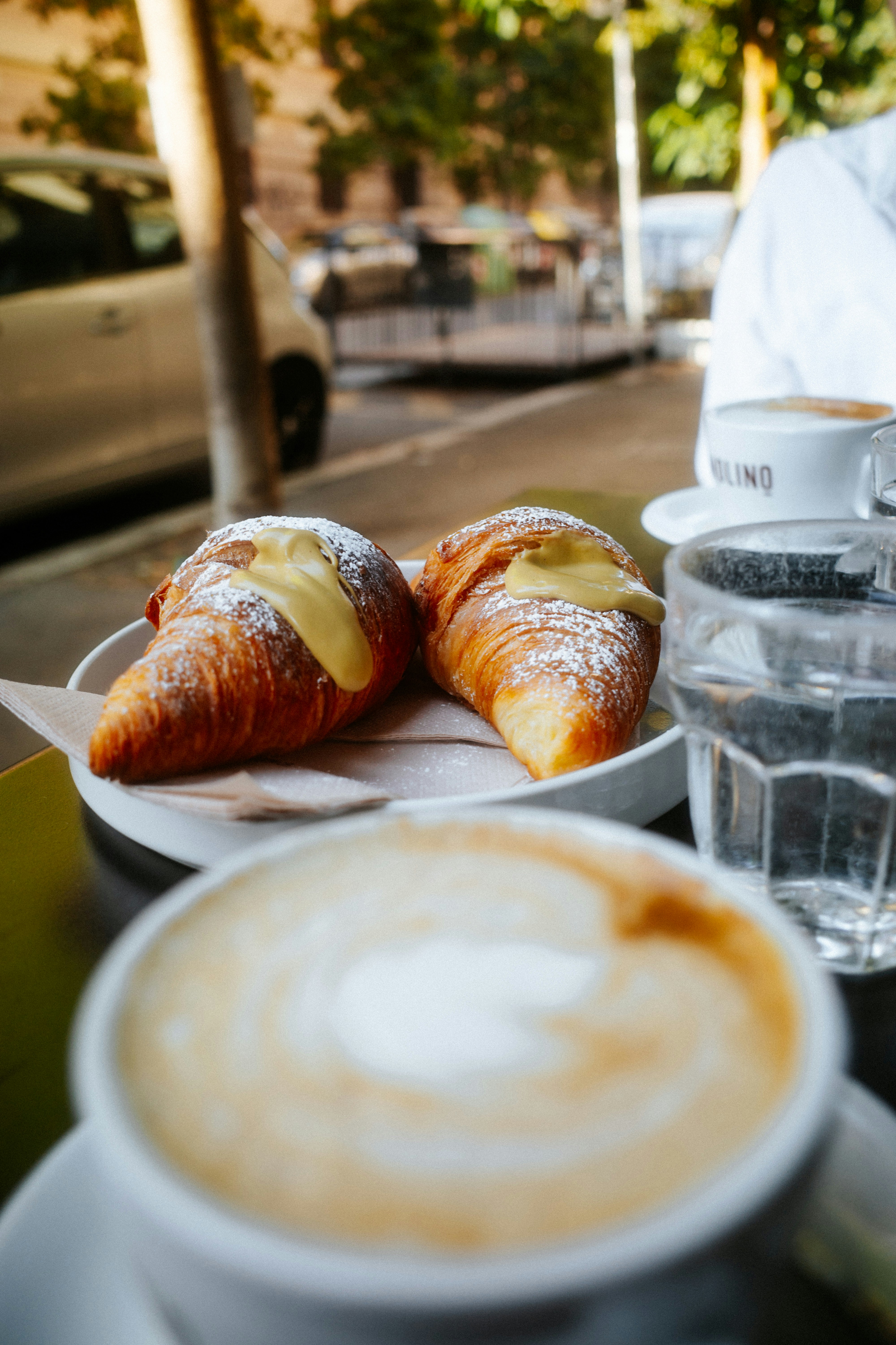 Crisp croissants with cream-touched centers sit on a plate beside a frothy coffee, with a cafe scene and glassware in the background.