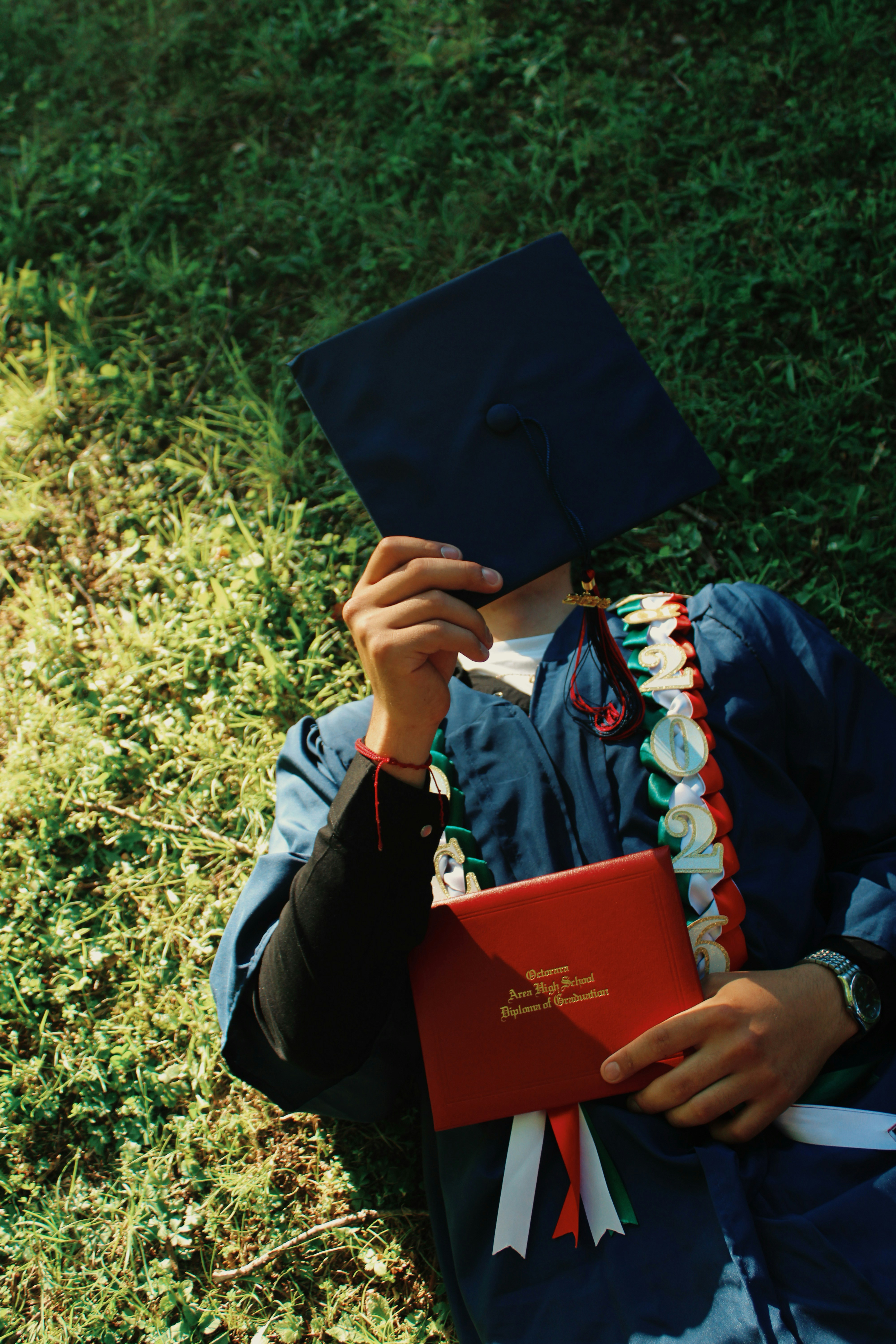 Graduate in cap and gown holds a red diploma, standing against sunlit grass with a ceremonial lei; the focus is on the celebratory moment and the hidden face adds intrigue.