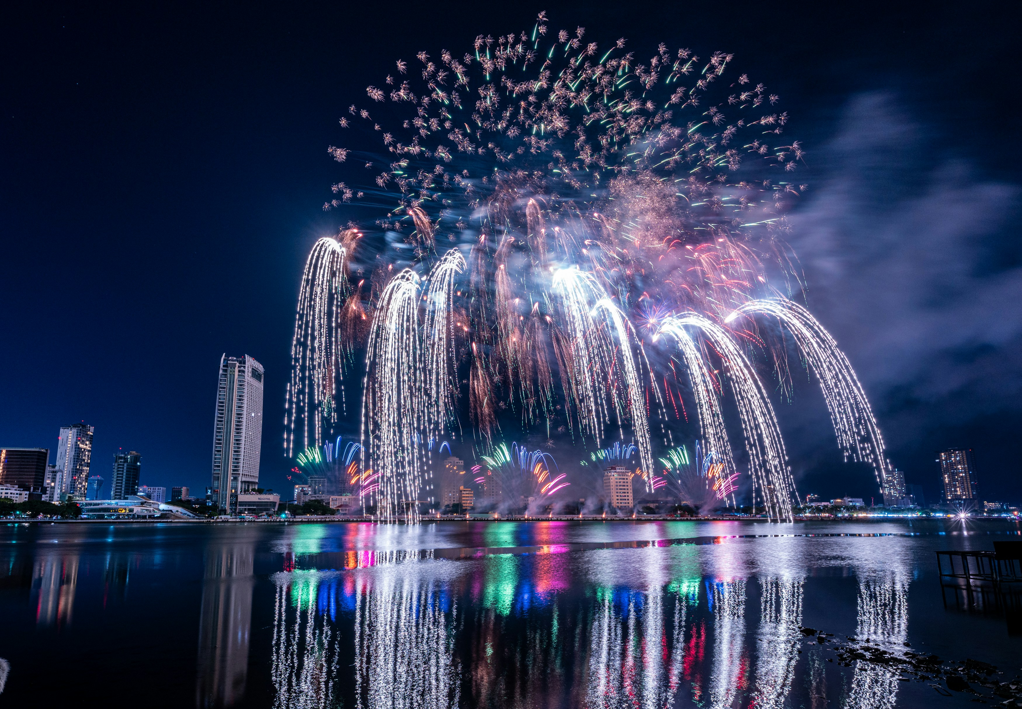 Fireworks explode over a city waterfront at night, mirrored in the calm water with illuminated buildings in the background. This is a photograph capturing vibrant colorful trails and reflections.