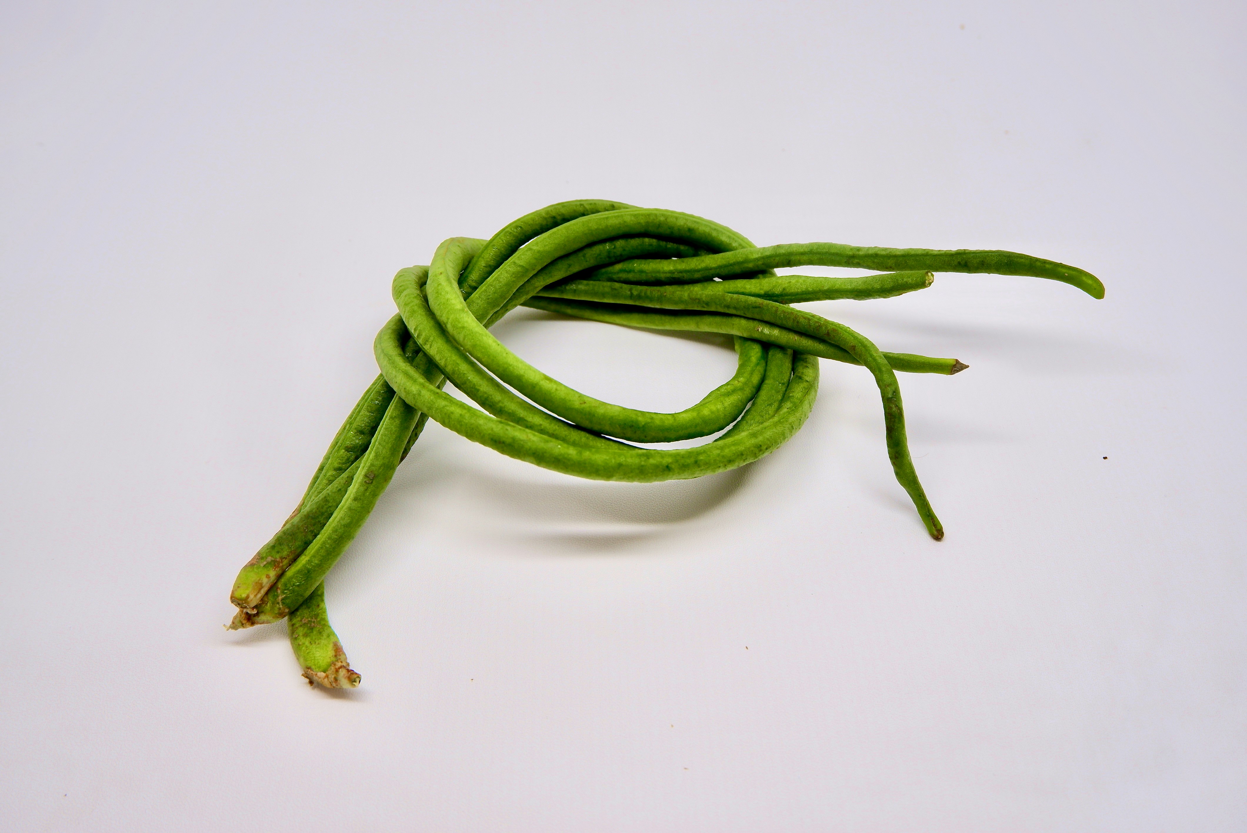 Fresh Raw Yardlong Beans Coiled on White. A studio close-up shot of fresh, vibrant green yardlong beans artfully coiled and arranged on a clean white background. This minimalist commercial image emphasizes the natural length, flexibility, and crisp texture of the raw vegetable, making it perfect for concepts related to healthy organic nutrition, vegetarian cooking, agricultural harvest, and authentic Asian culinary ingredients.