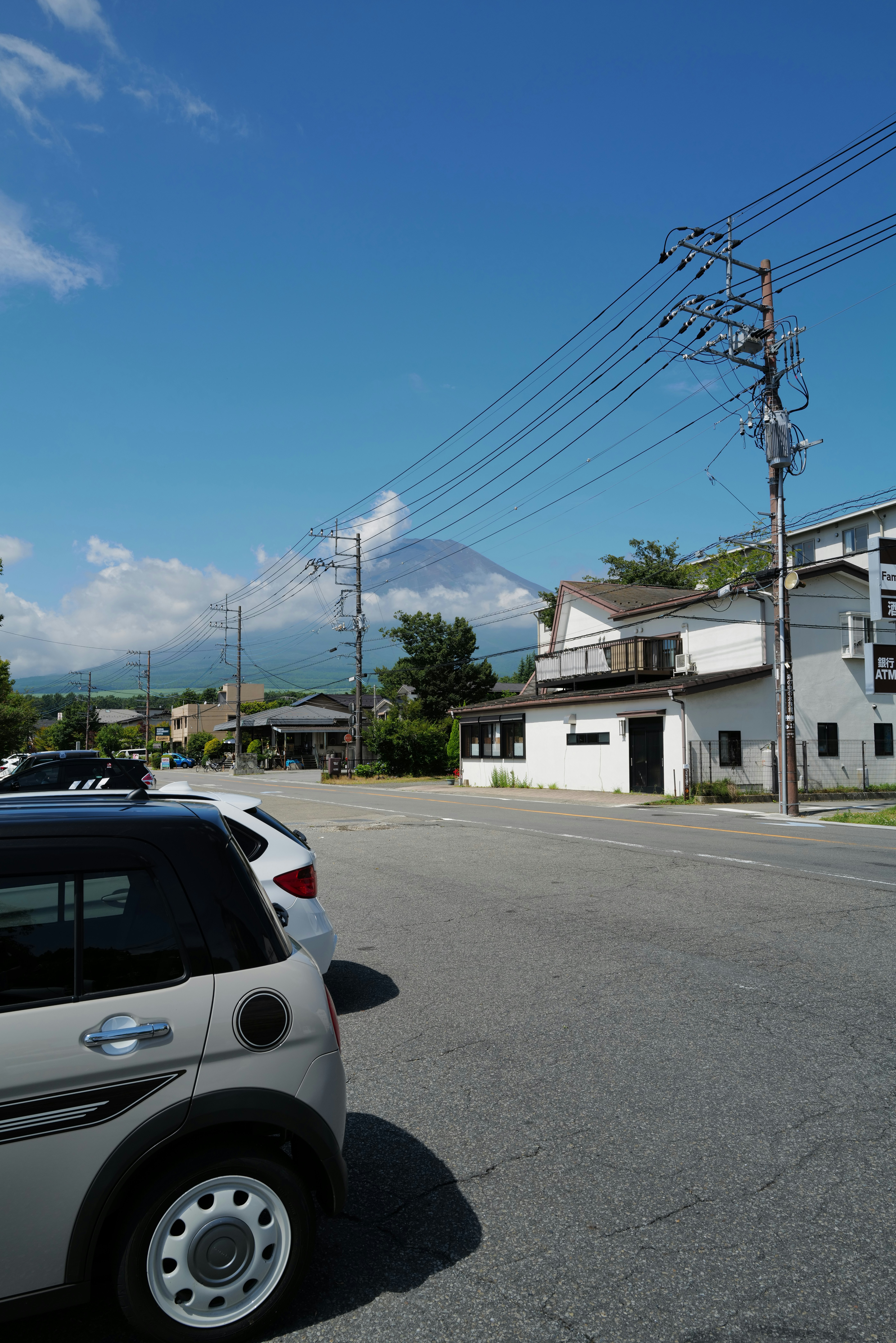 Wide street scene with parked cars in the foreground and a distant volcano-like hill under a clear sky; utility poles run along the road, while suburban buildings line the right. A calm daylight scene with strong perspective toward the distant peak.