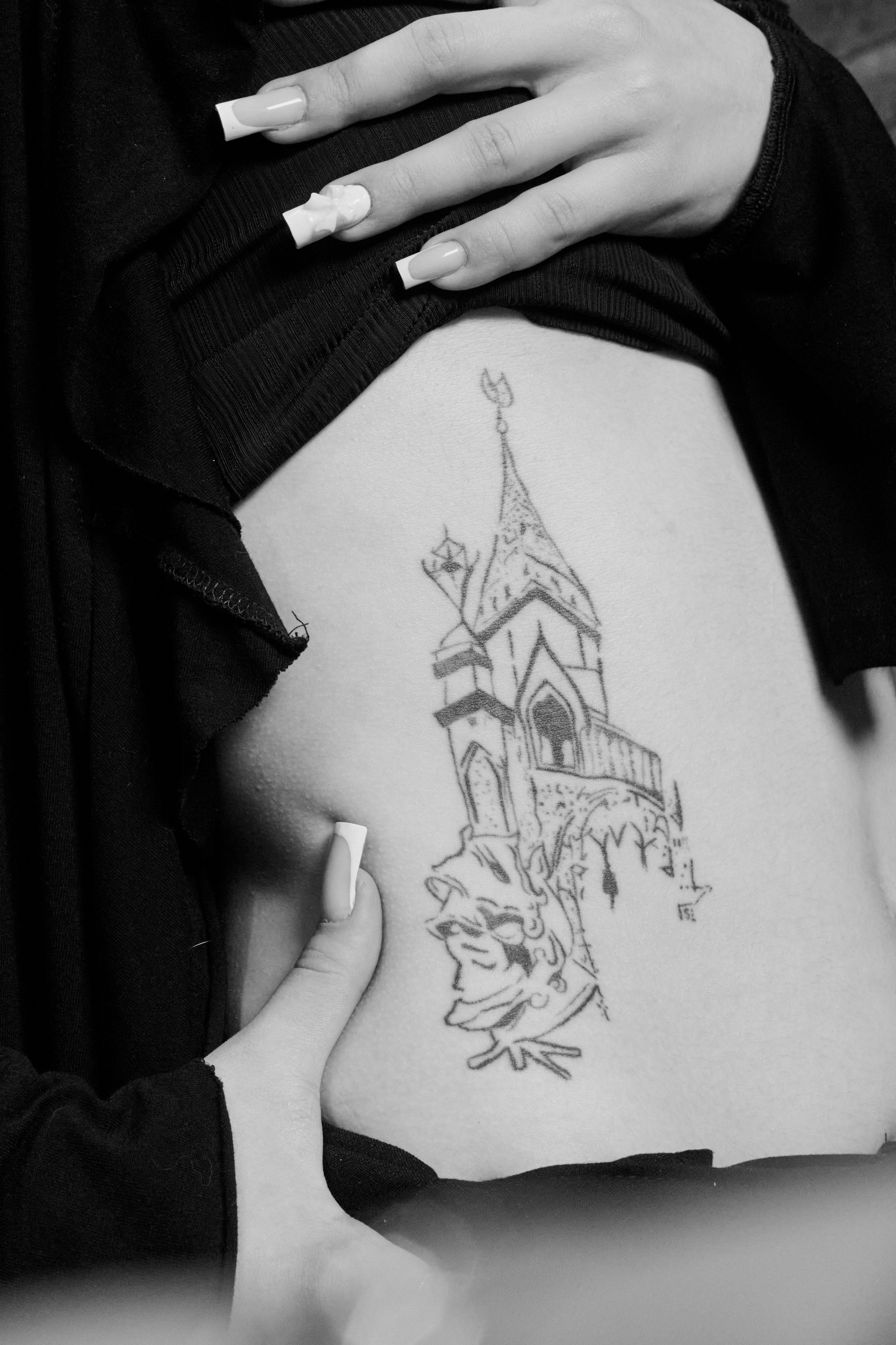 Tattooed architectural church motif along the side of a torso, framed by hands lifting a cropped shirt; grayscale portrait emphasizing linework and skin texture.