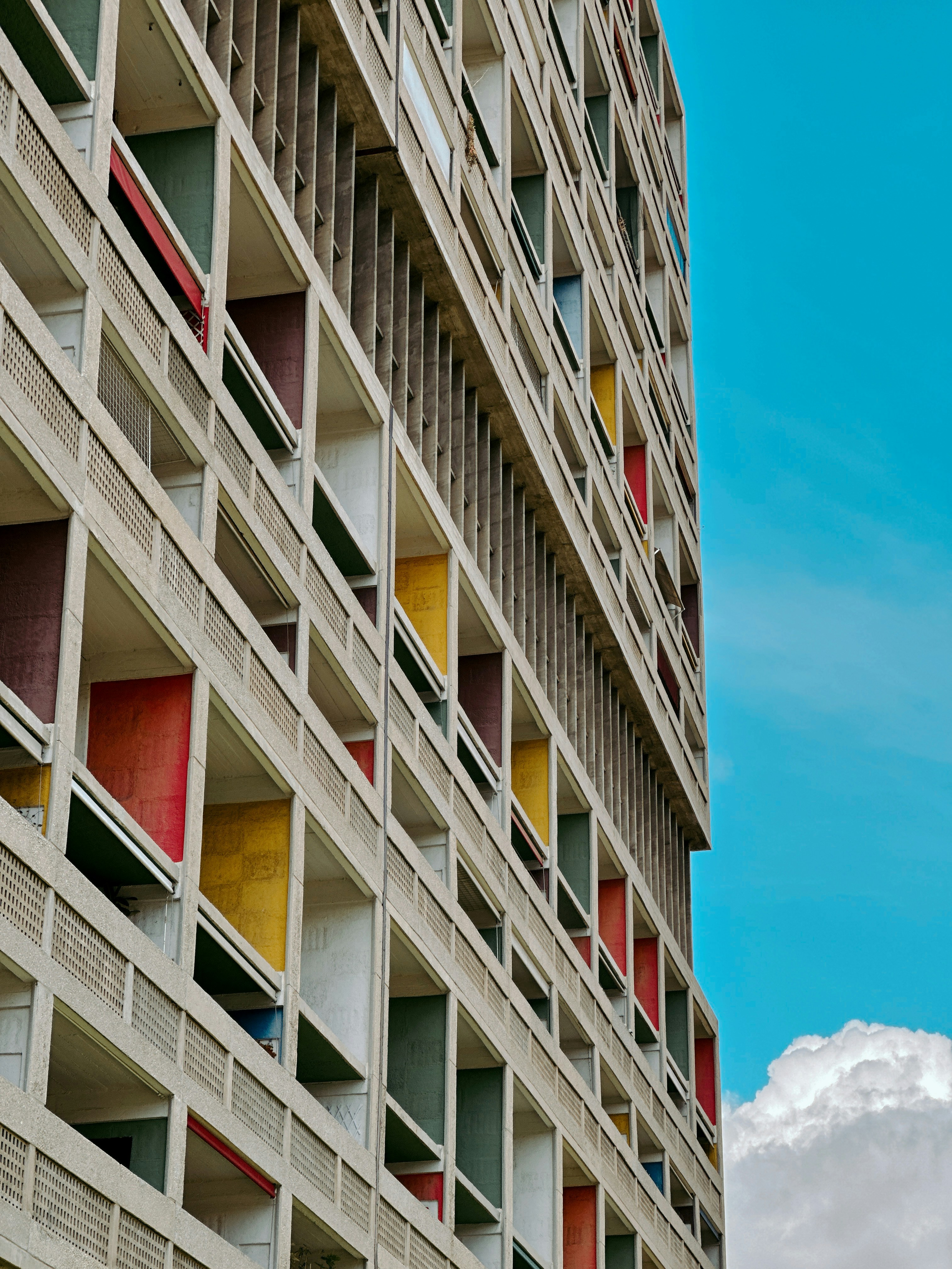 Textured apartment facade with multicolored balcony panels extending diagonally against a blue sky. Architectural rhythm and color blocks give a dynamic, abstract feel to the scene.