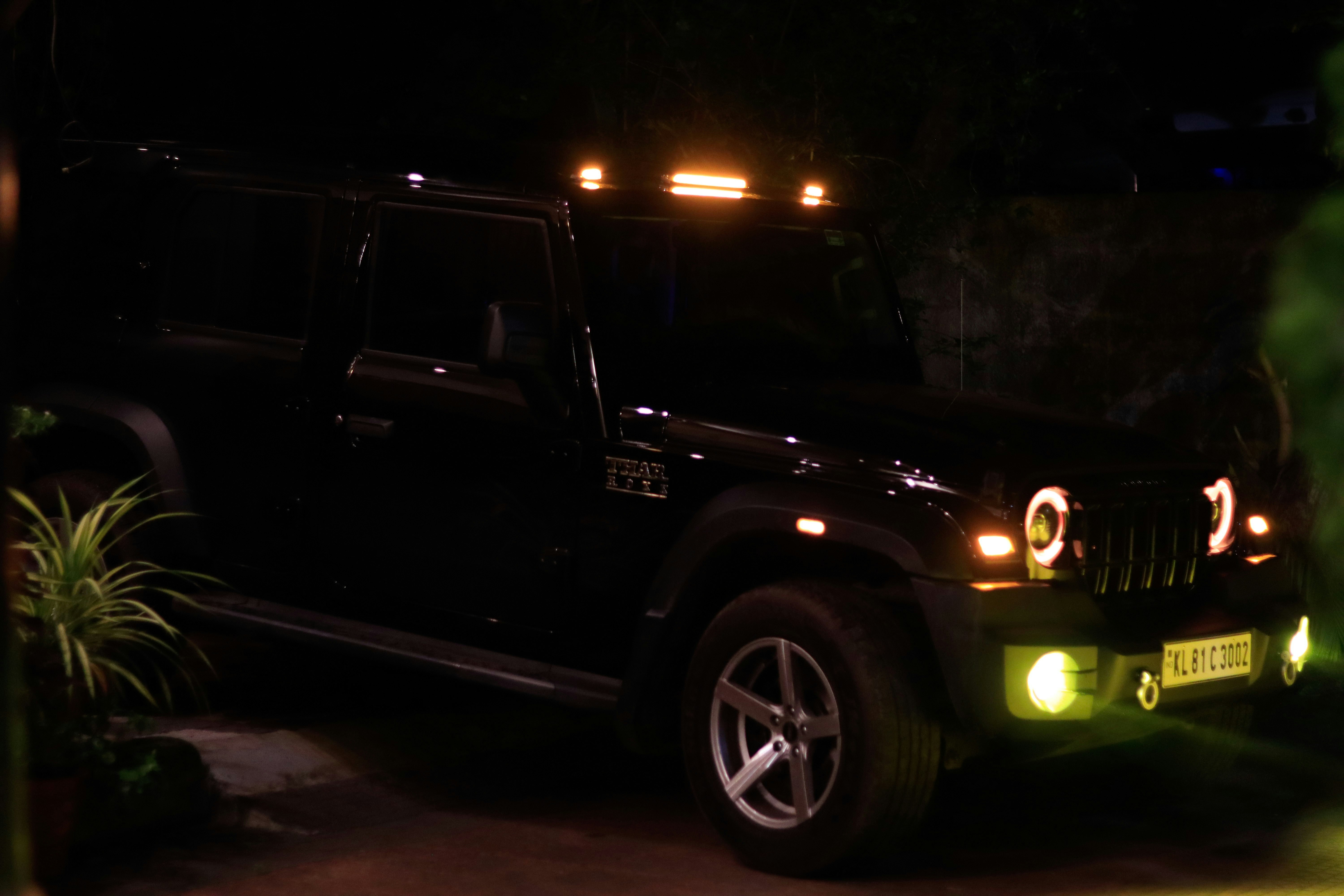 Dark SUV parked at night with bright front fog lights and rooftop lights, subtle highlights on body panels; a car in a driveway scene.