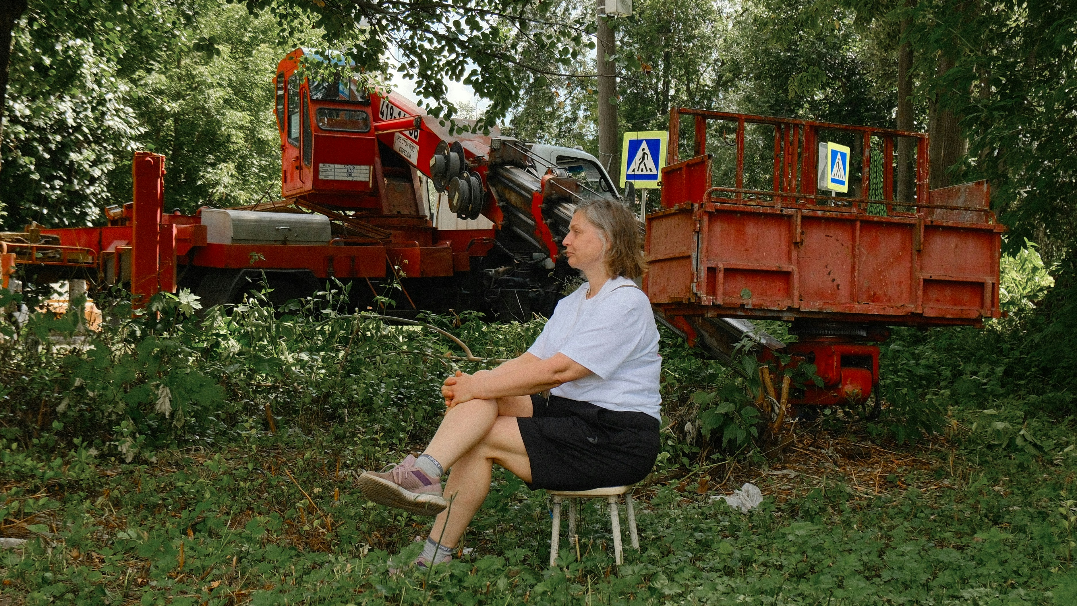 Woman in casual white tee sits on a stool beside a red industrial machine under leafy trees