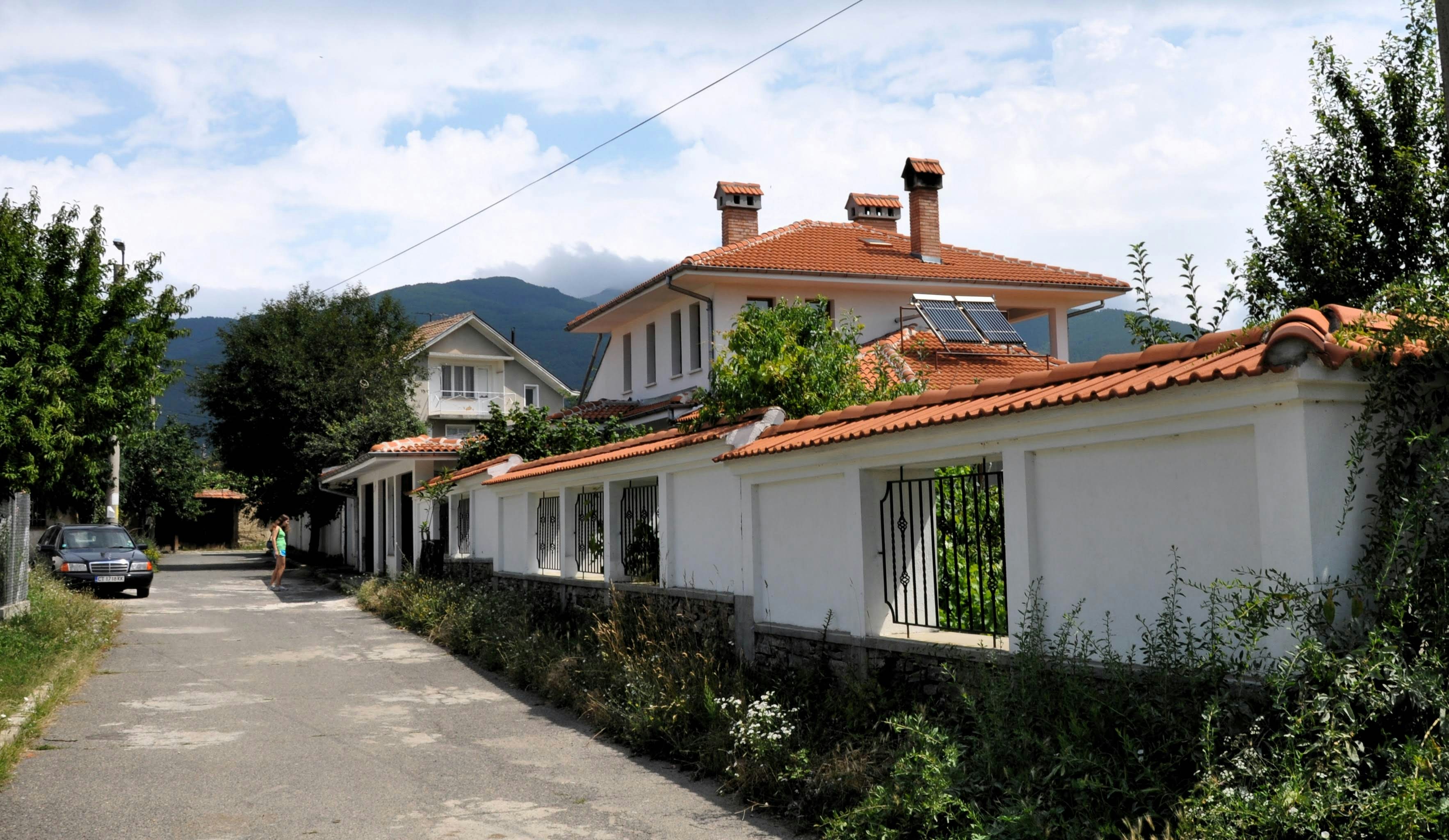 Sunlit street of white-walled houses with red-tiled roofs and a distant hillside, showcasing a quiet residential scene.