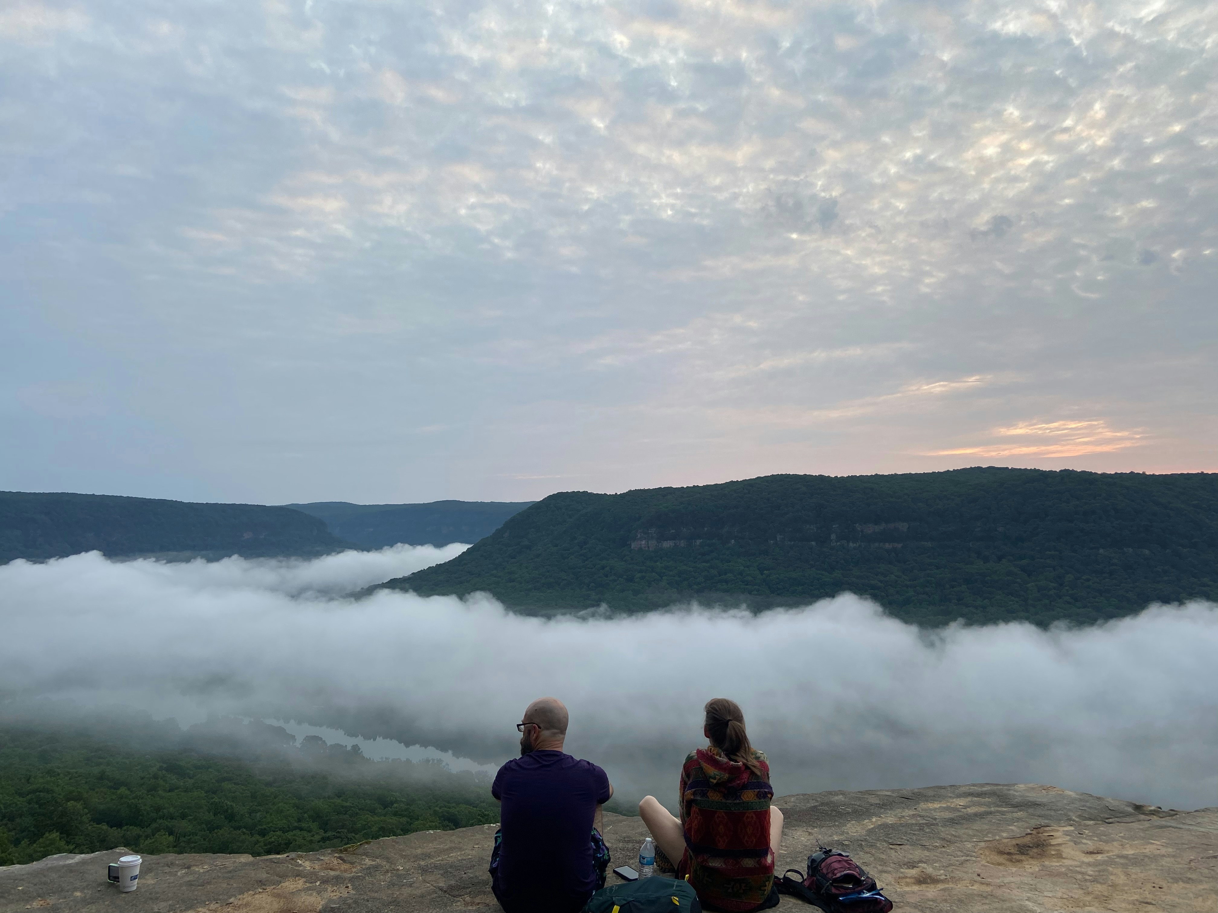 Two hikers on a cliff watching a sea of clouds over a forested gorge