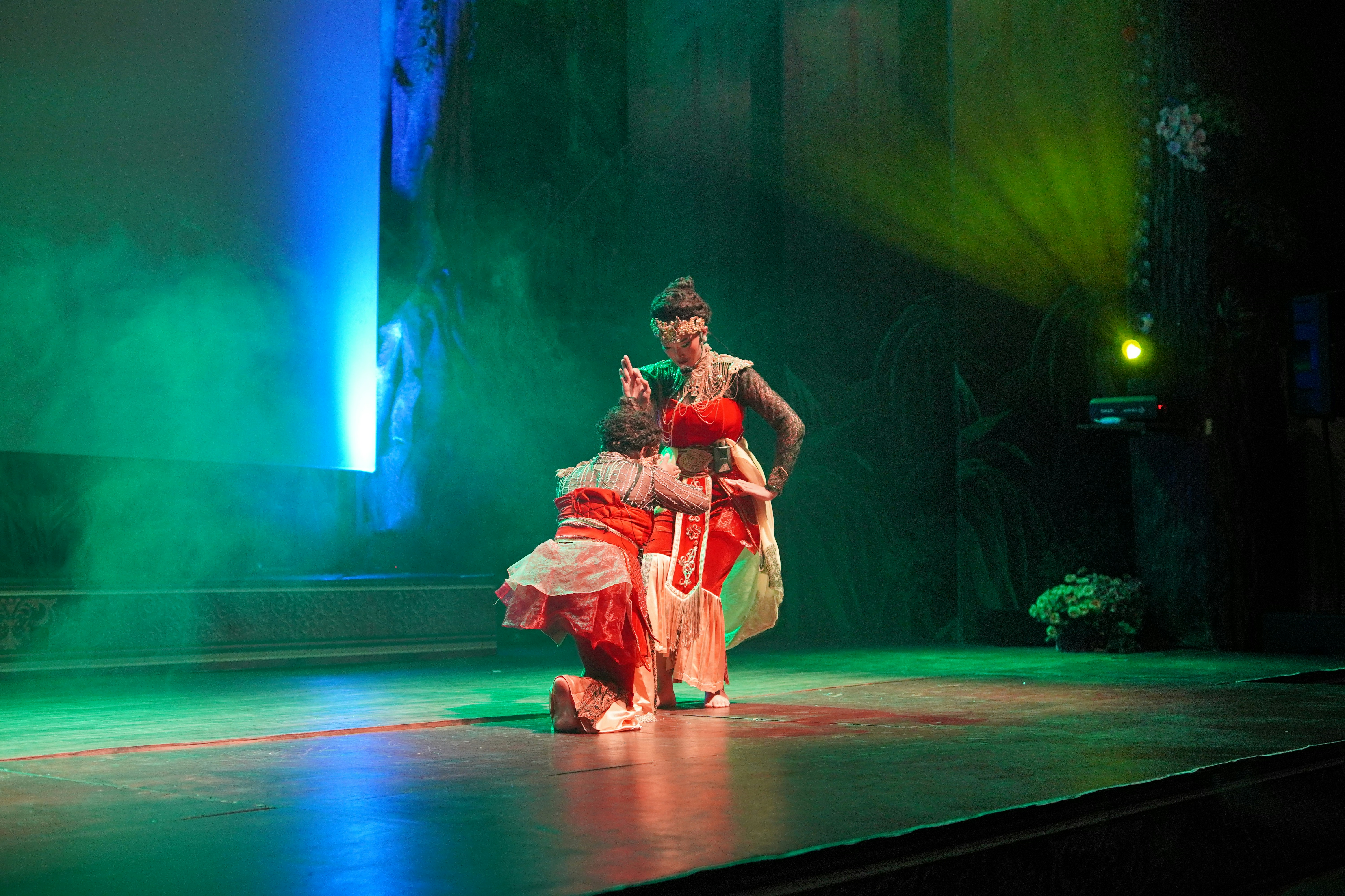 Energetic dance duo in vibrant red costumes performs on a foggy, color-lit stage with green and blue hues. The action centers on the lead dancer mid-mip, creating a sense of motion and theatrical composition.