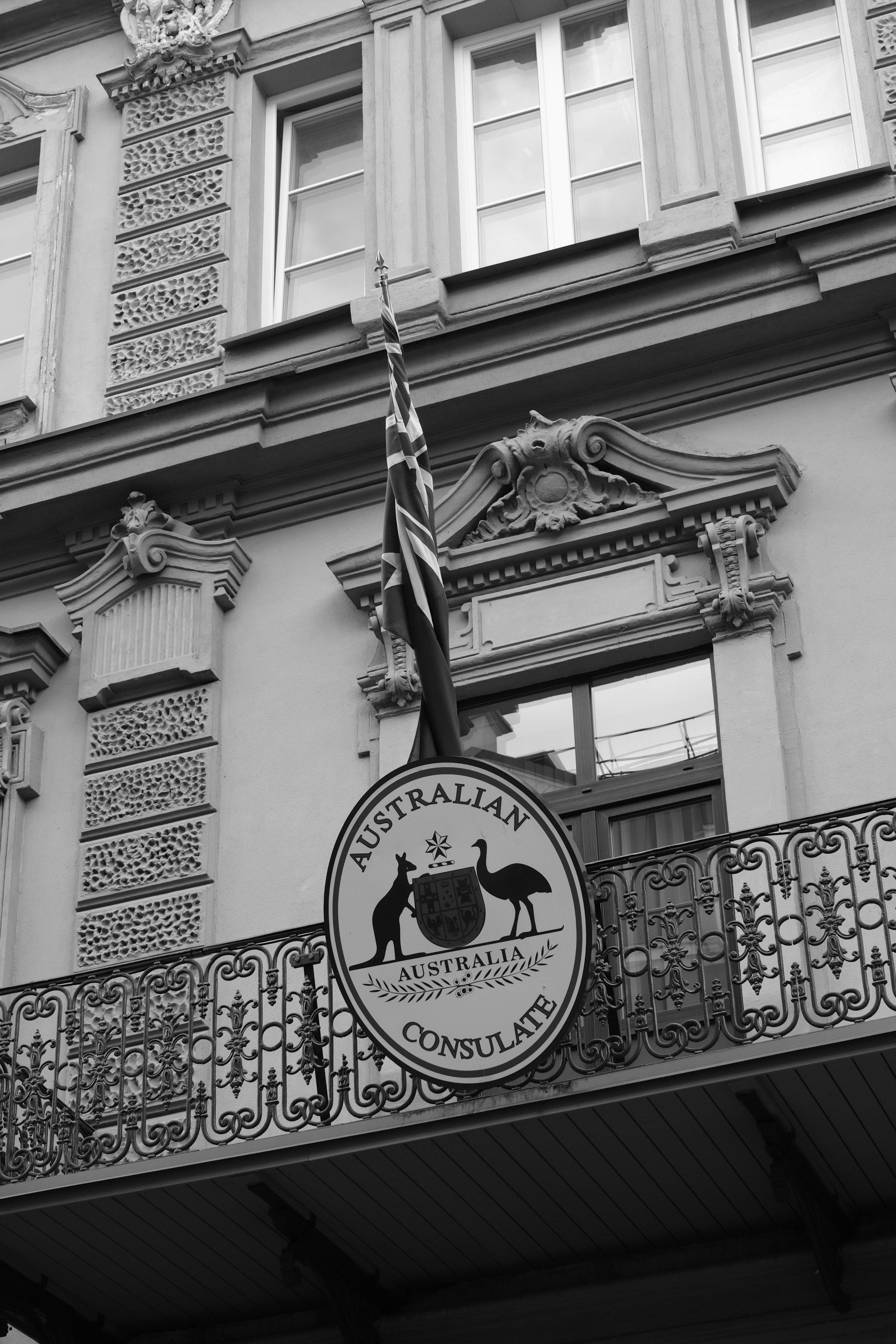 Black-and-white facade of the Australian Consulate building featuring a hanging emblem and ornate iron balcony railing.
