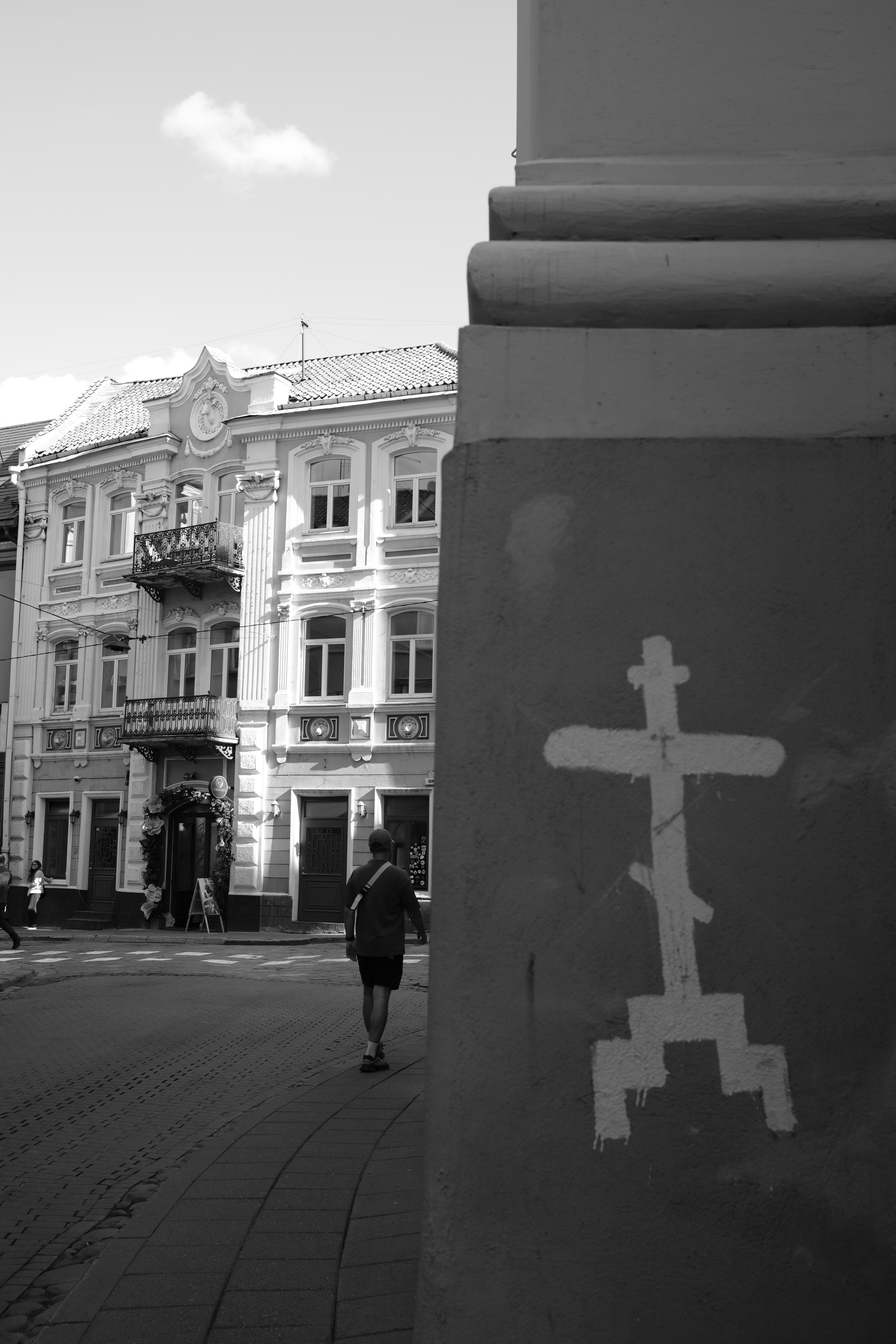 Monochrome street scene featuring a pedestrian walking past a large concrete column adorned with a white cross graffiti, and classic European buildings in the background.