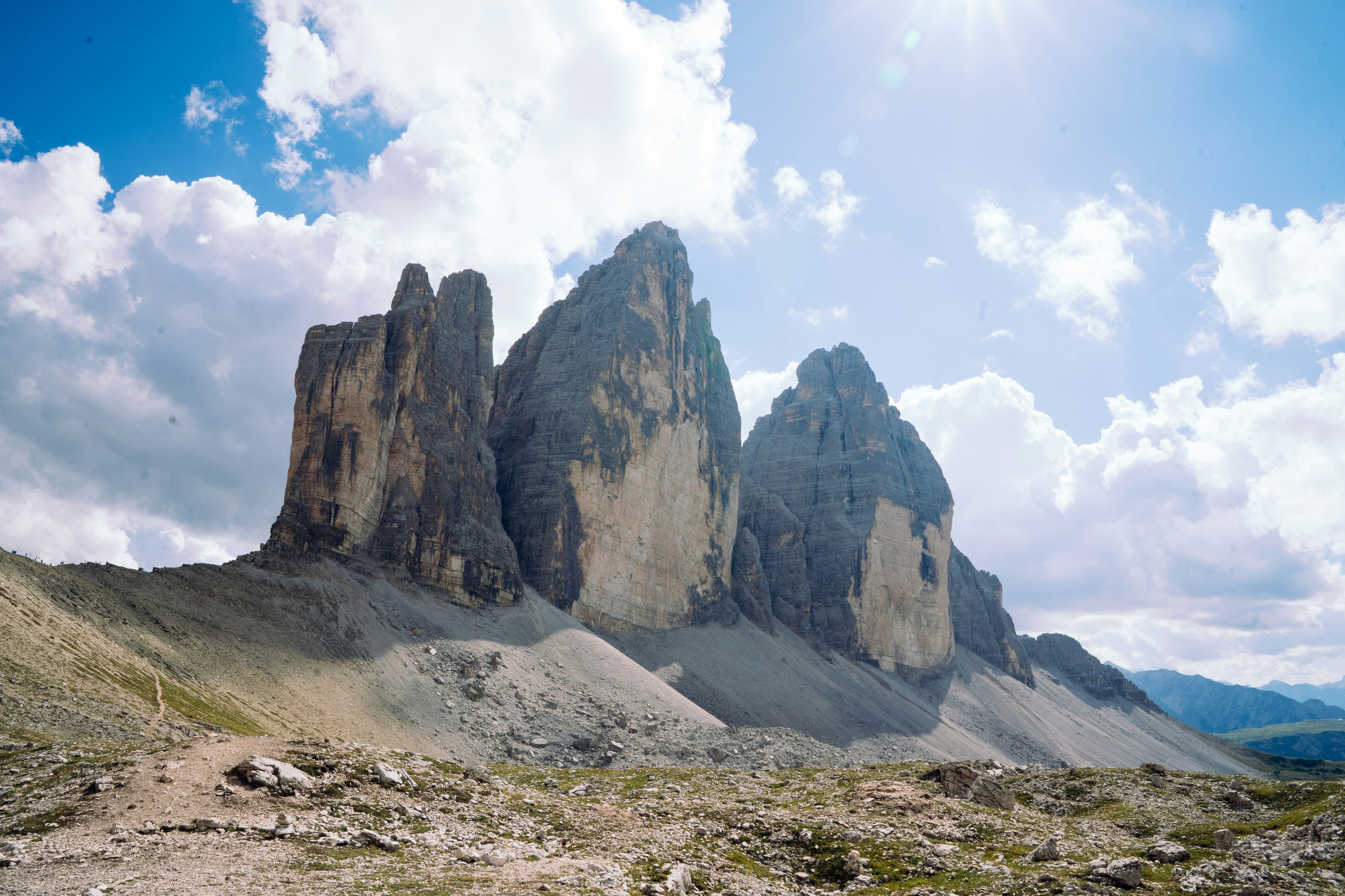 Three towering rock formations rise above a barren slope under a bright sky, with rugged scree in the foreground and distant ridges fading into the horizon. A landscape shot capturing the scale and textures of the mountain trio.